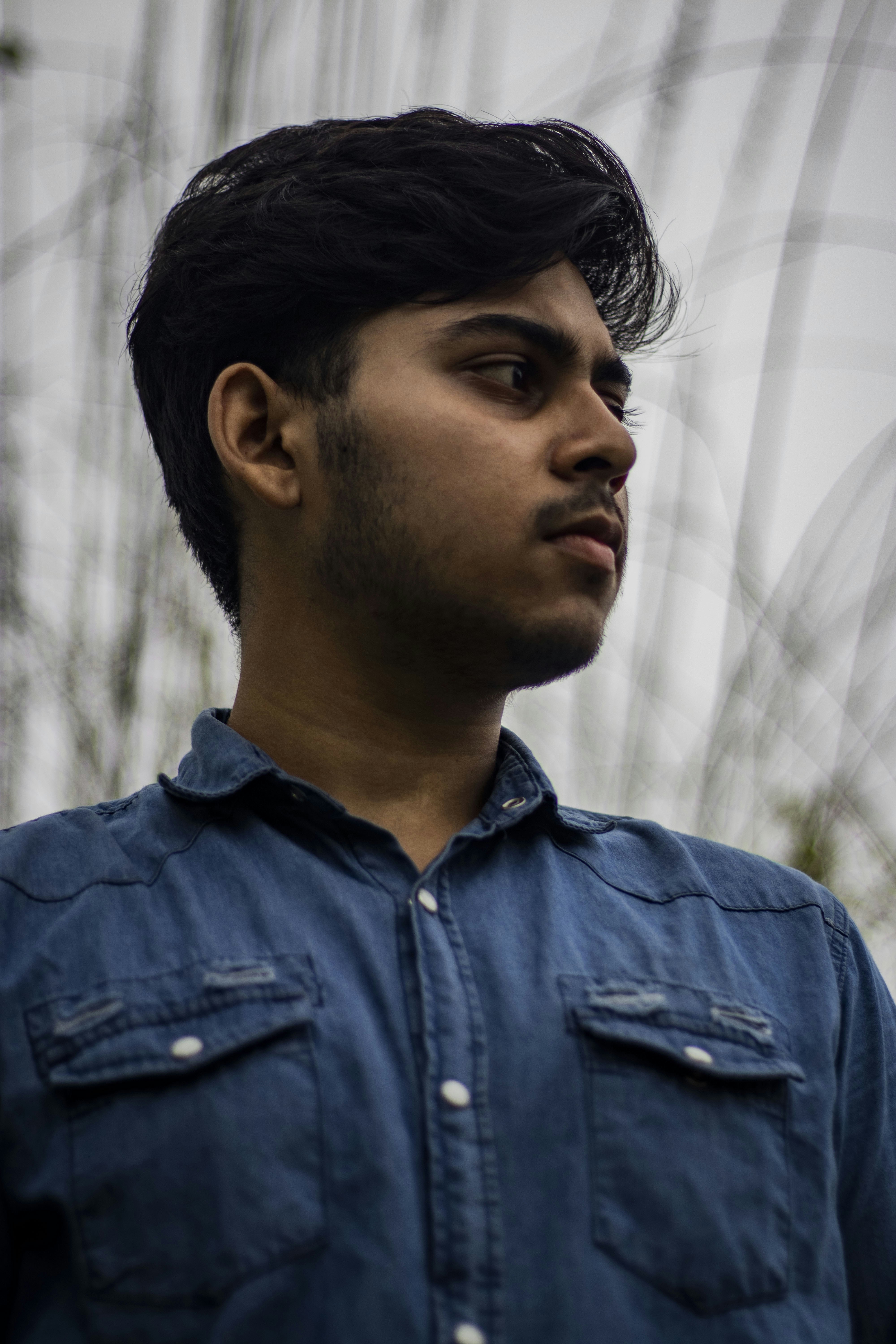 Young man in a denim shirt gazing thoughtfully, surrounded by tall grasses under a soft, overcast sky.