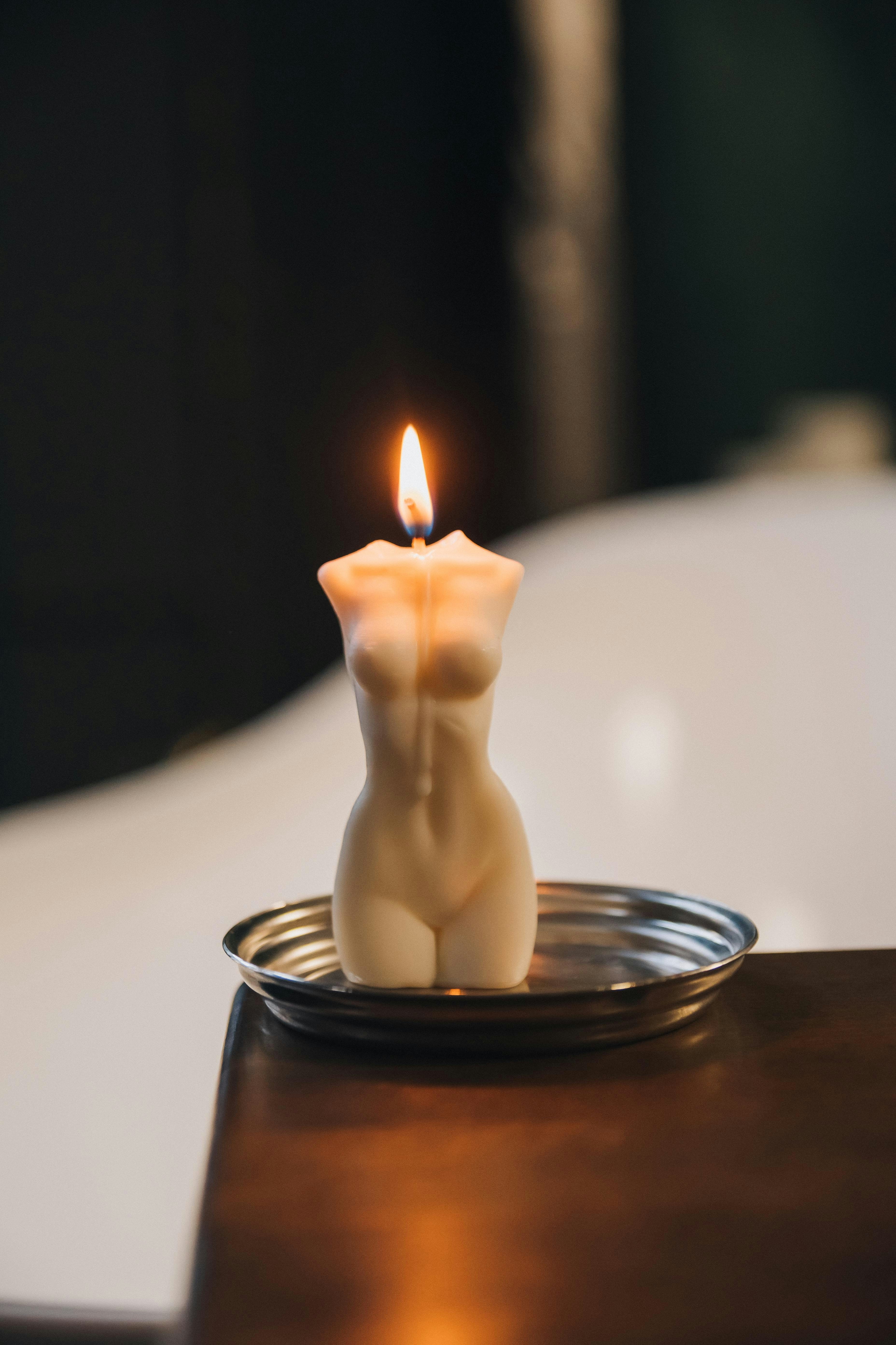 A sculptural candle shaped like a torso, elegantly lit with a flickering flame, resting in a metal dish against a softly blurred background.