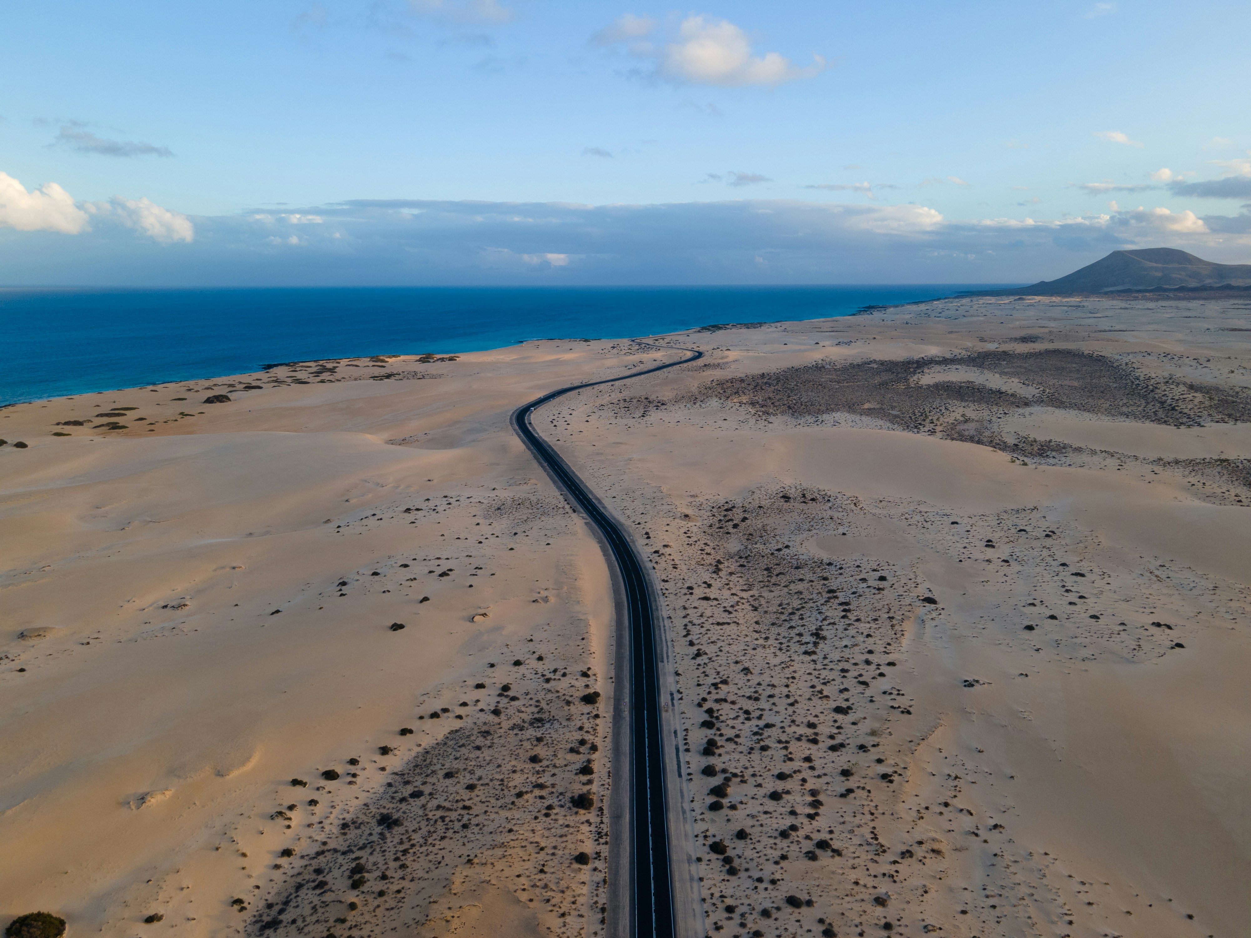 A winding road cuts through vast sandy terrain, leading towards the tranquil blue ocean under a partly cloudy sky.