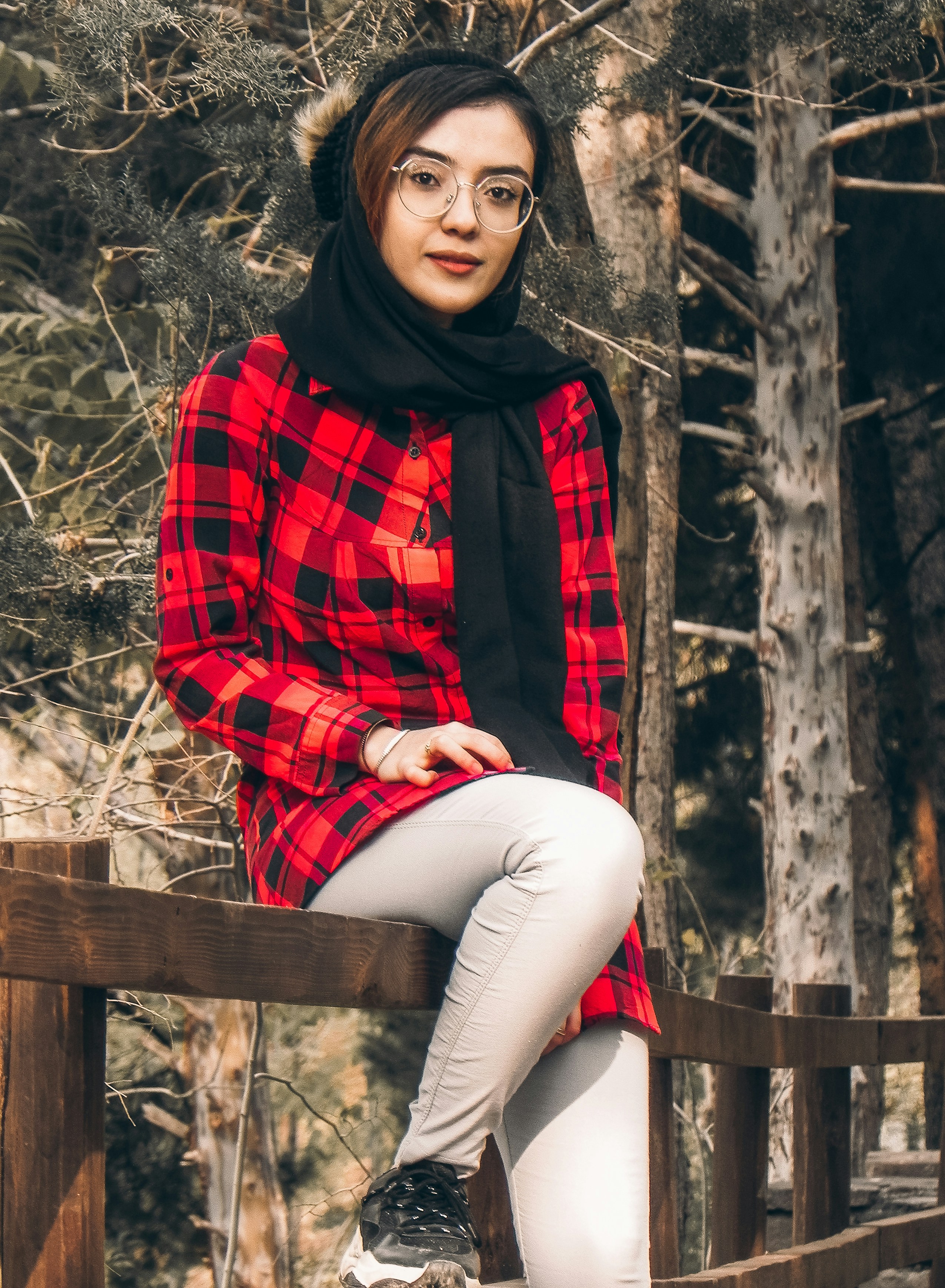 Young woman in a red plaid shirt and black scarf sitting on a wooden railing in a forest setting, surrounded by trees.