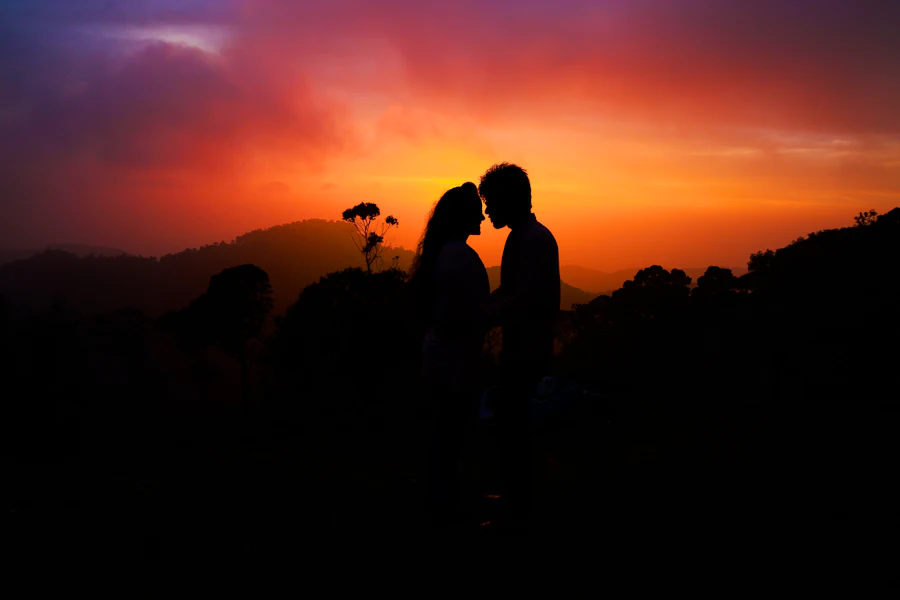 A silhouetted couple stands close together against a vivid orange and purple sunset, with dark hills and trees framing the scene.