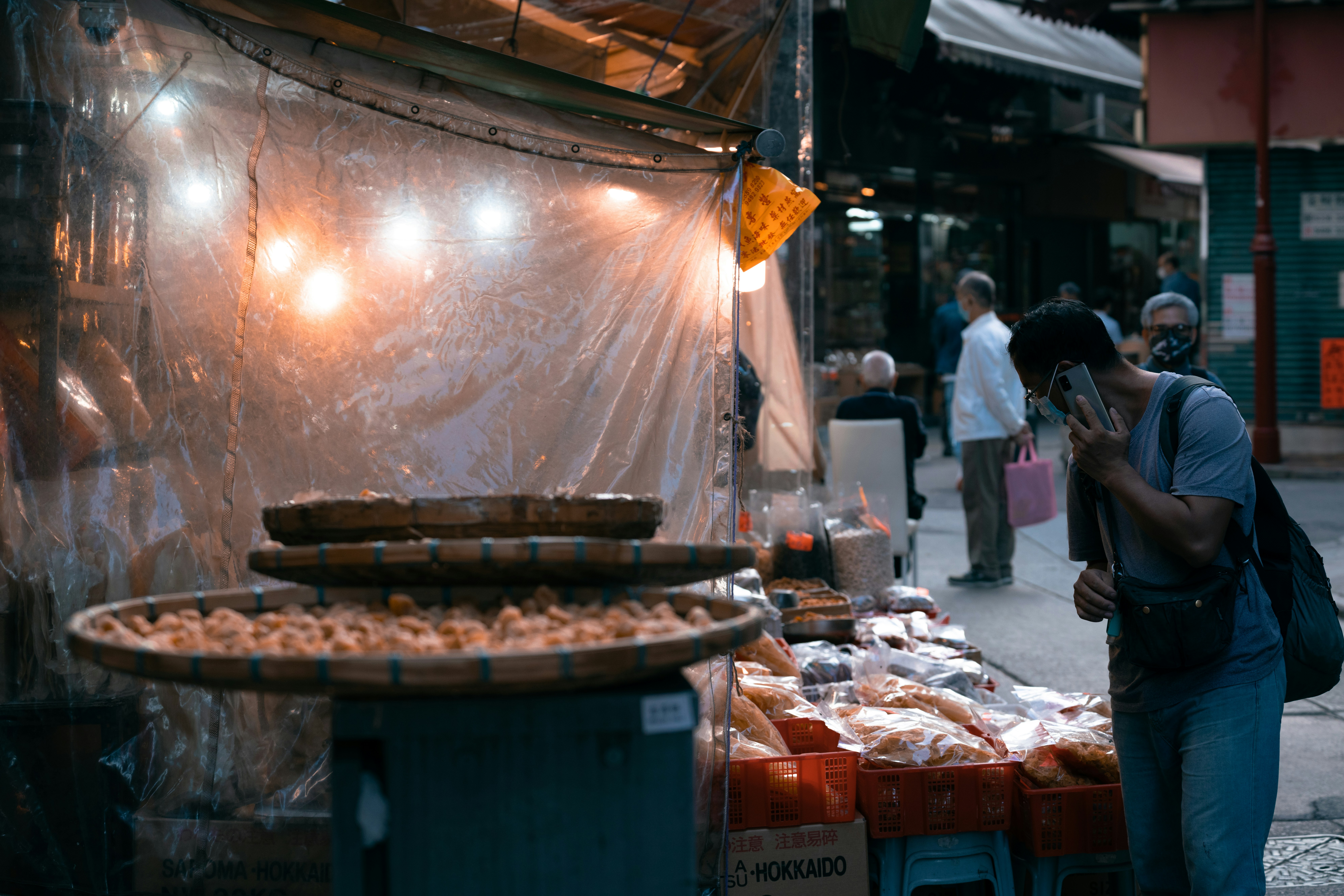 A vendor stall illuminated by warm lights contrasts with the twilight ambiance, while a figure stands in contemplation amidst a vibrant array of goods.