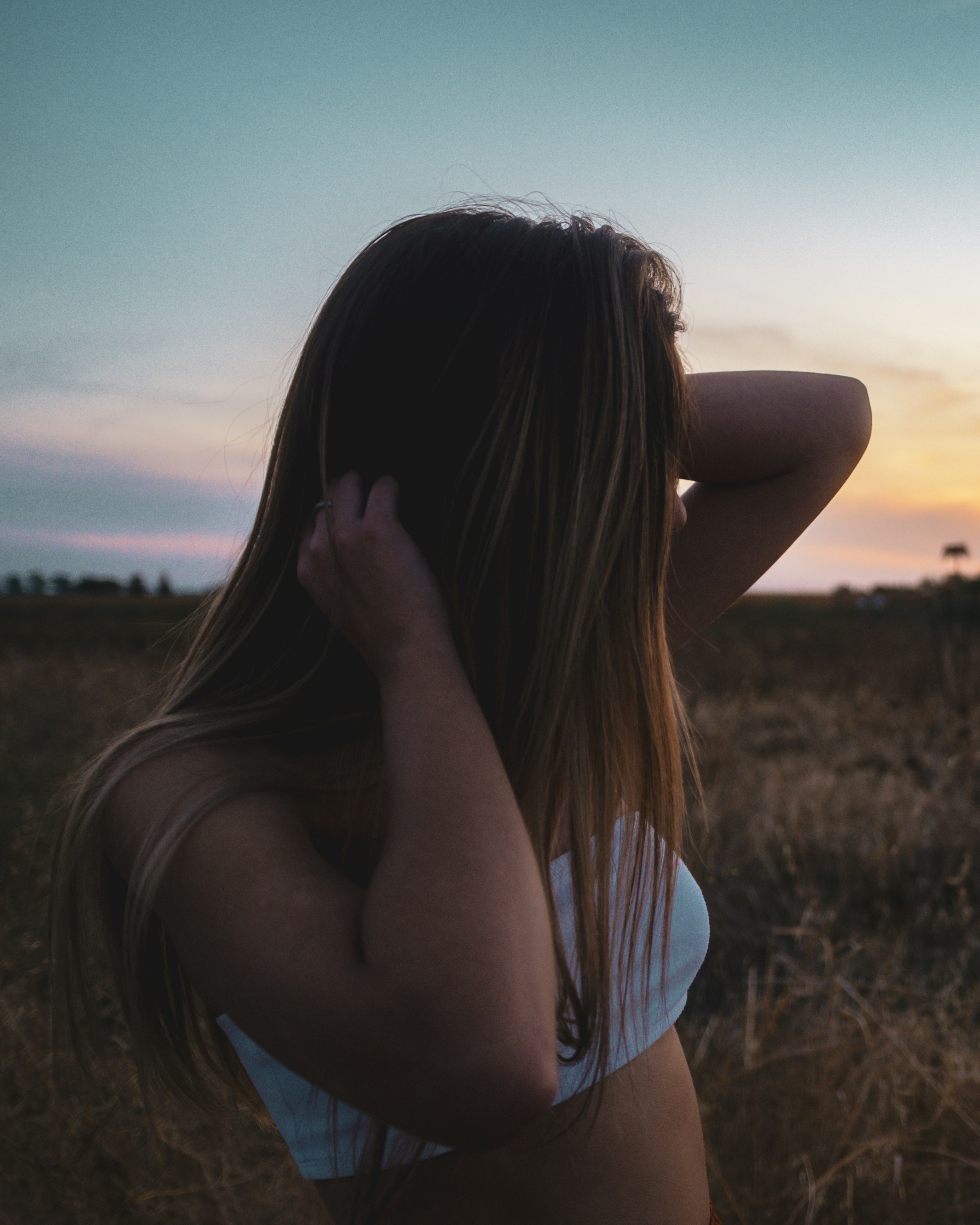 Silhouette of a woman with long hair, gently holding her arm behind her head against a twilight sky. The scene captures a serene moment in a grassy landscape.