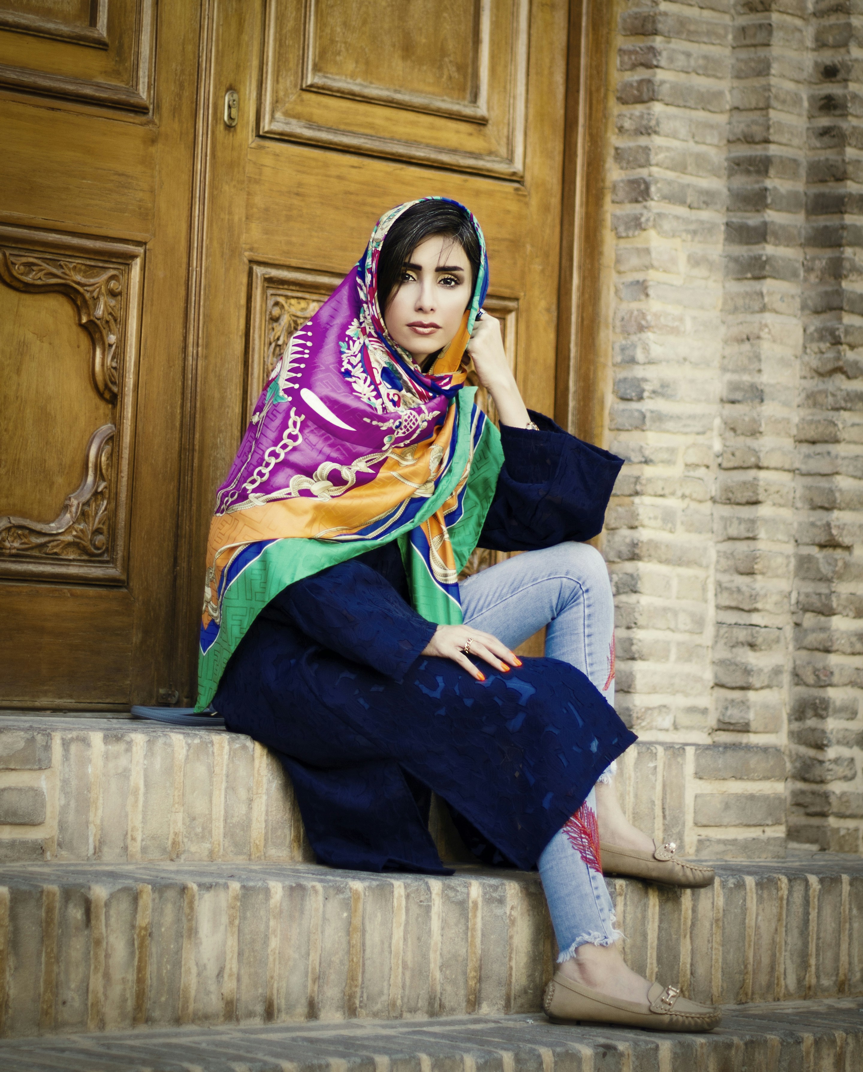 A woman seated gracefully on stone steps, adorned in a colorful scarf and traditional attire, with a wooden door and brick wall as a backdrop.