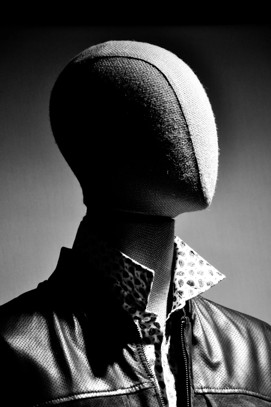 Black-and-white portrait of a faceless mannequin or dress form wearing a patterned collared shirt and jacket, lit from the side against a dark background. Strong contrast emphasizes the fabric textures and silhouette.