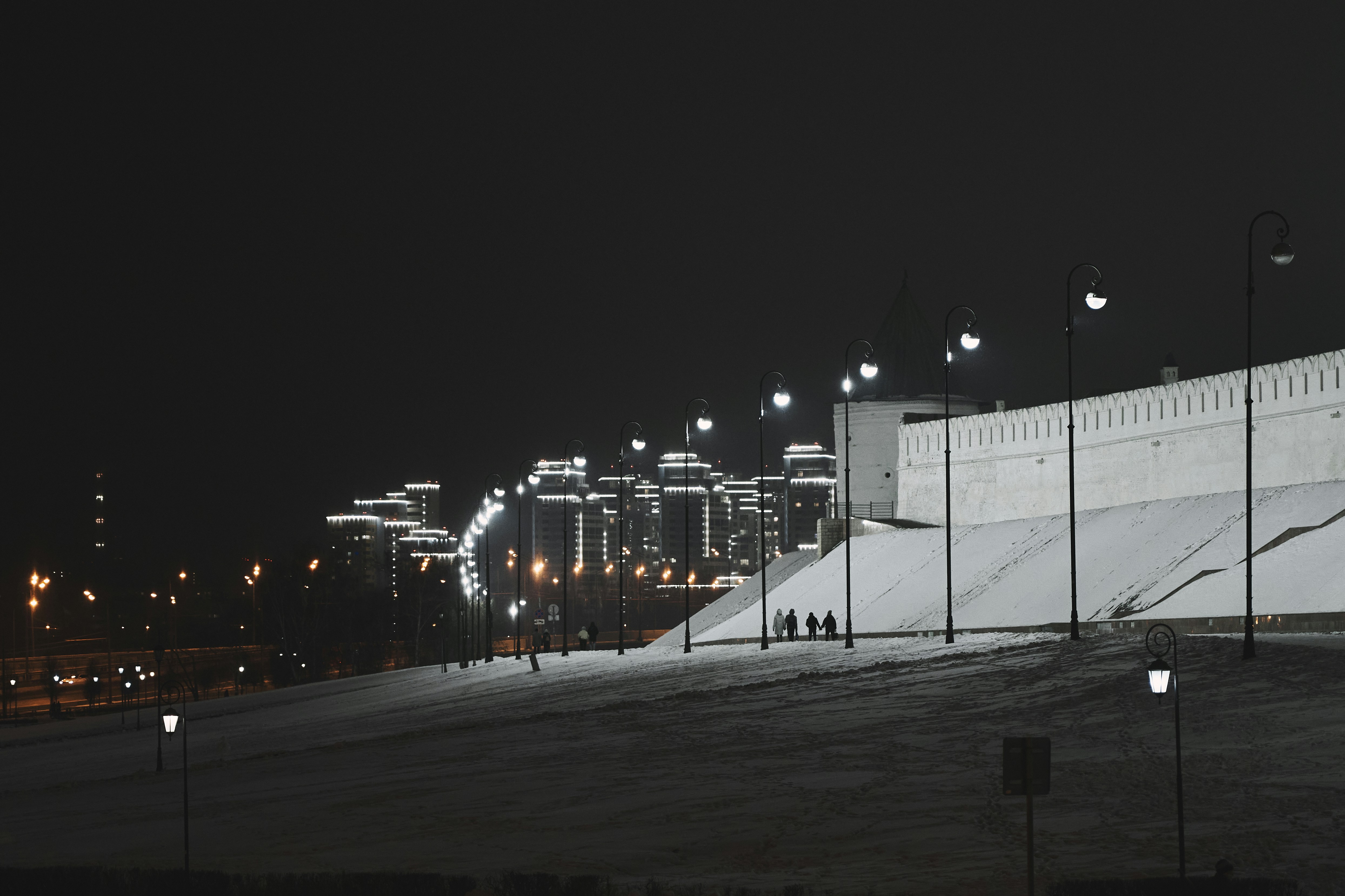 Snow-covered slope leading to a historic fortress wall, illuminated by street lamps, with modern city skyline in the background. 