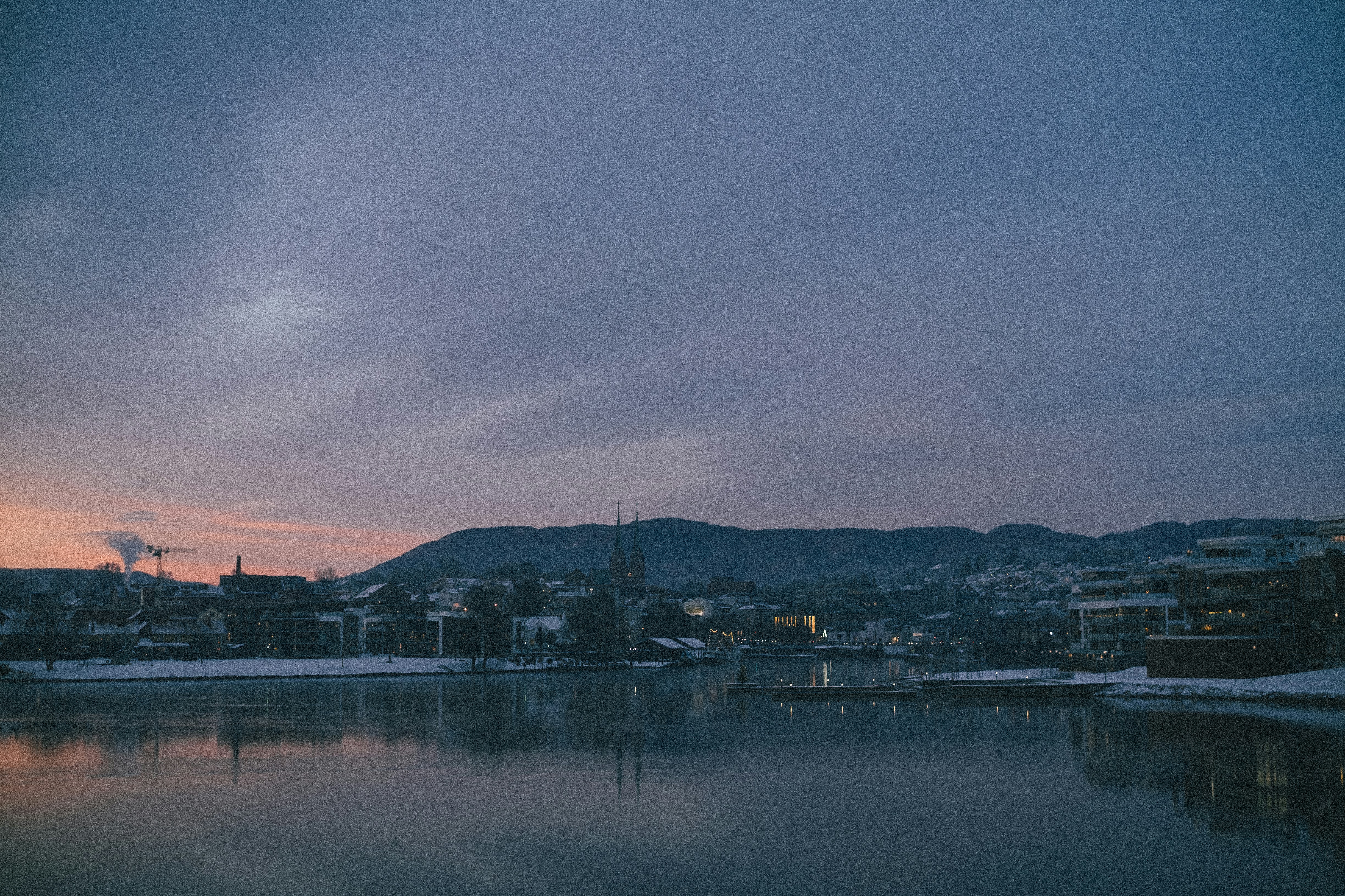 A tranquil lakeside scene at twilight, showcasing a quiet town silhouetted against a softly lit sky. The still water reflects the muted colors of dusk.