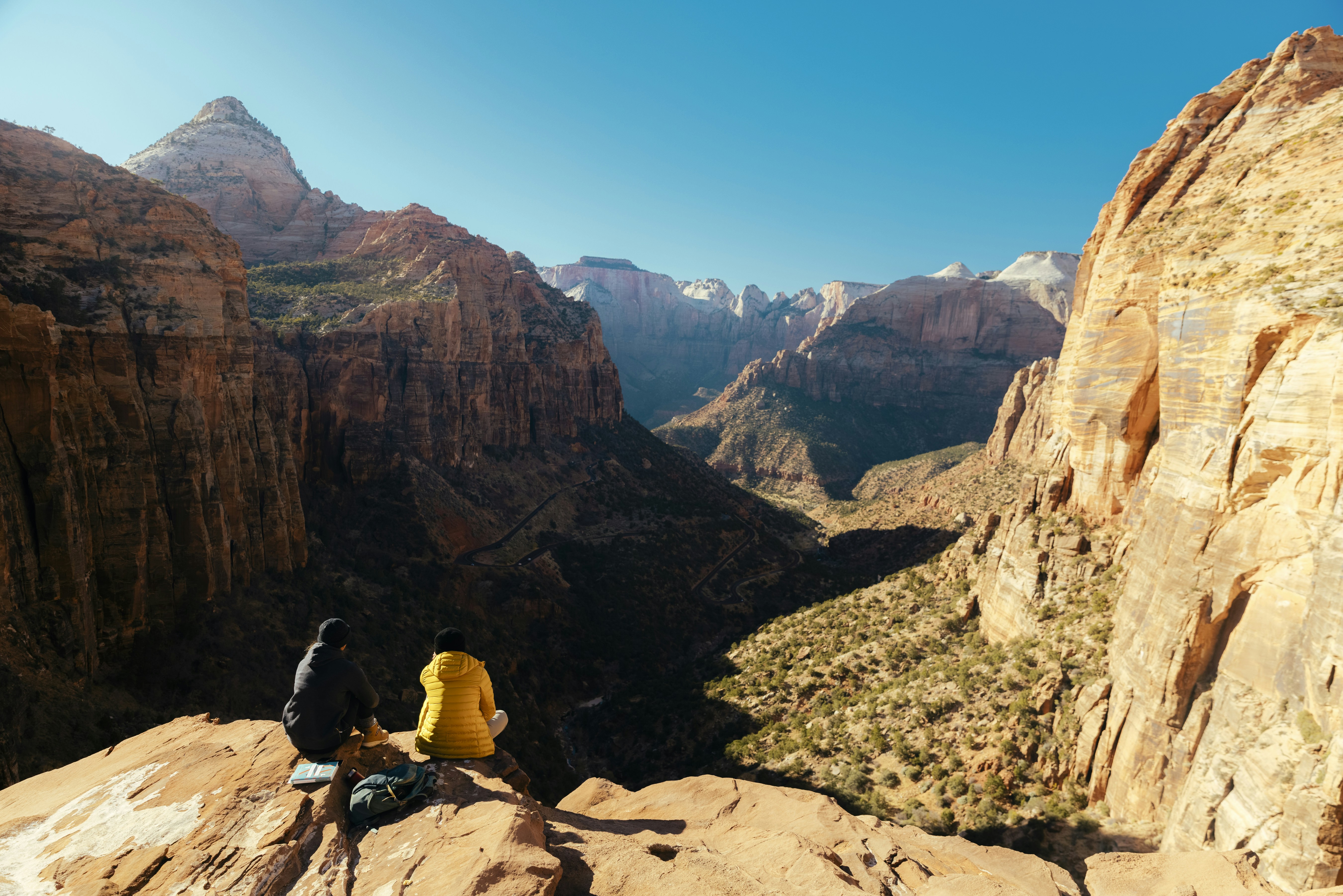 Two people seated on a sunlit rock overlooking a vast canyon under a clear blue sky.