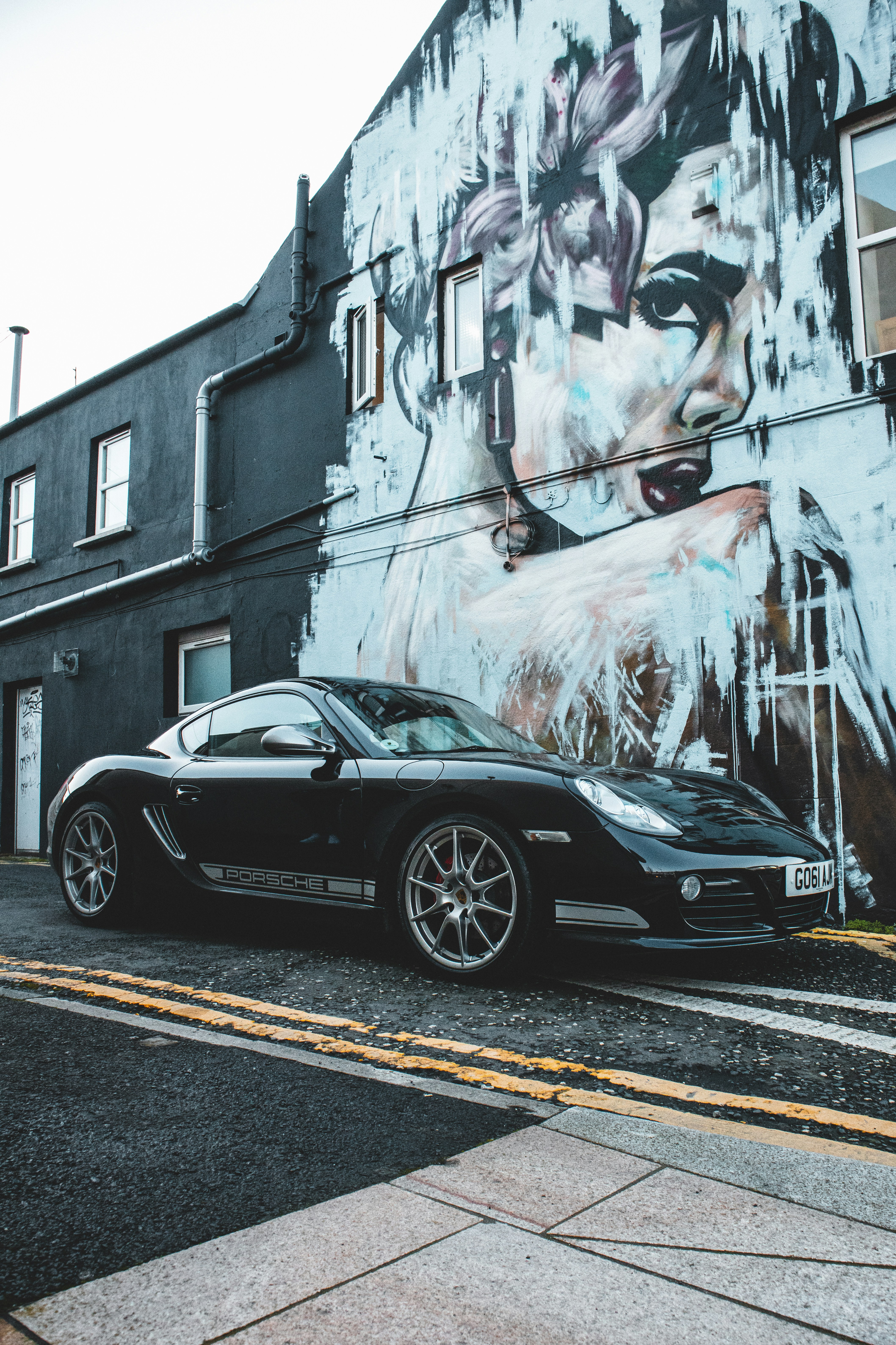 A sleek Porsche sports car parked beside a vibrant mural depicting a woman's face, showcasing the fusion of automotive design and urban artistry.
