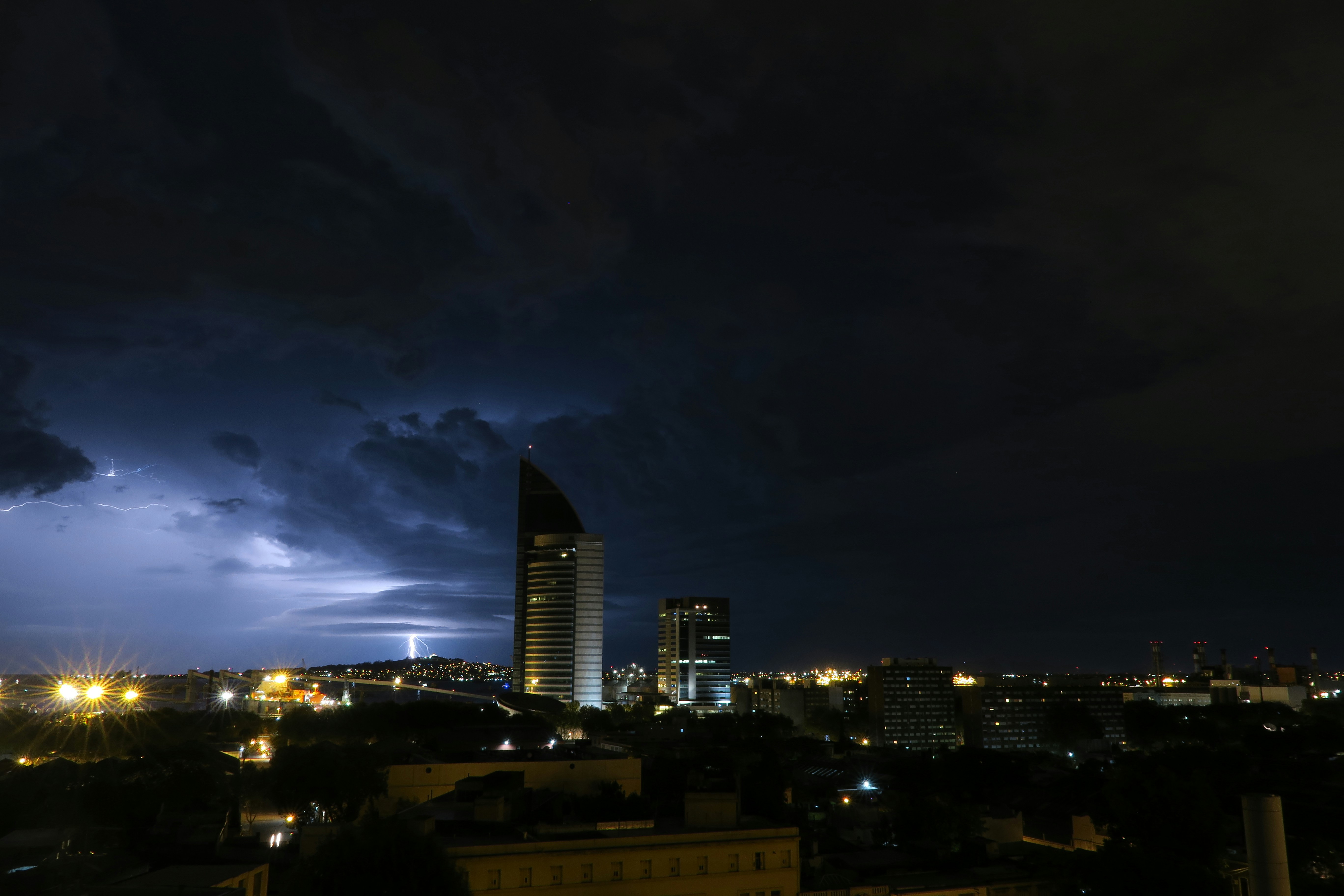 Dark storm clouds illuminated by lightning loom over a city skyline at night, showcasing modern architecture against a dramatic backdrop.