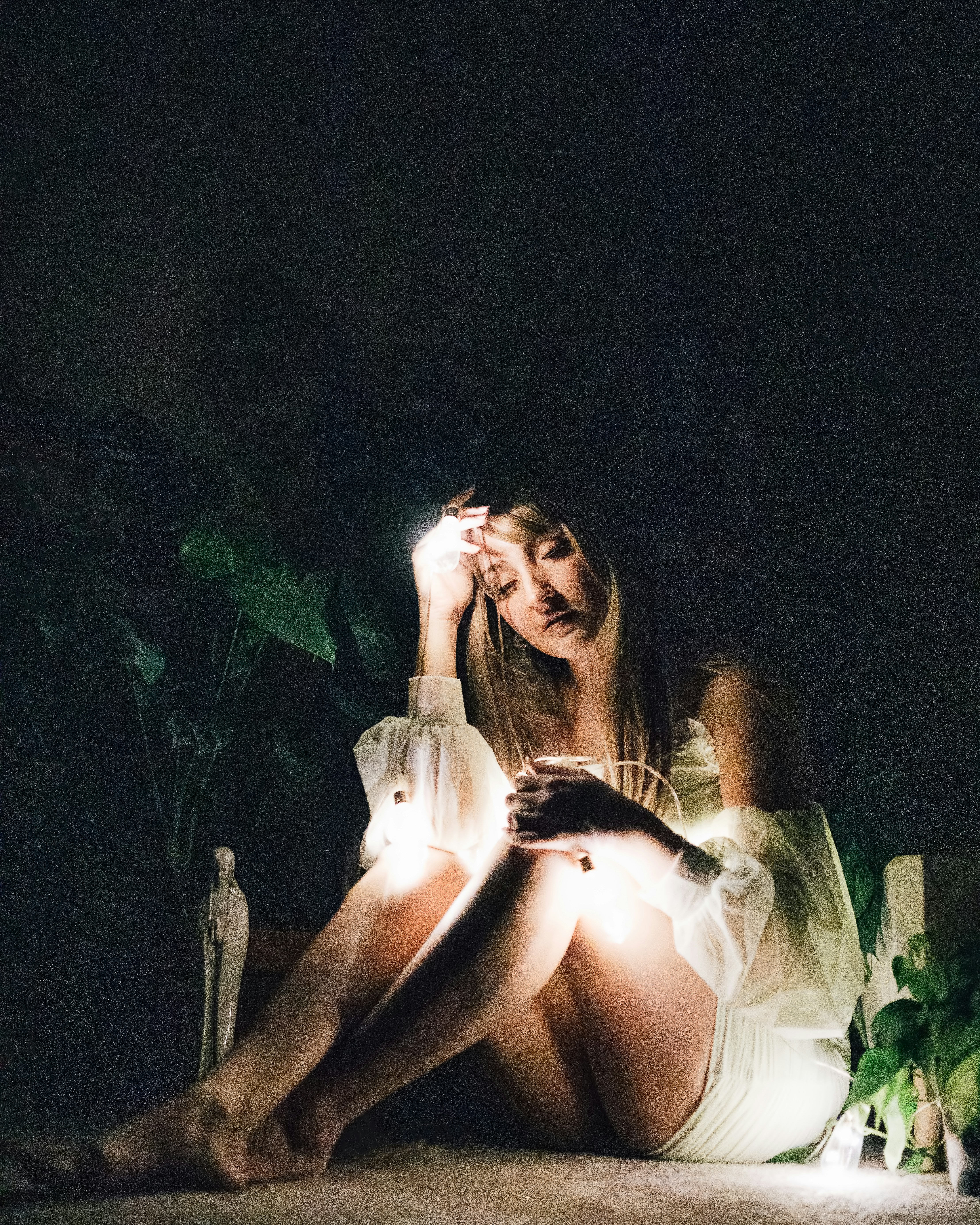 A contemplative figure seated in soft light, surrounded by lush greenery, creating a serene atmosphere. The play of light enhances the mood of introspection.