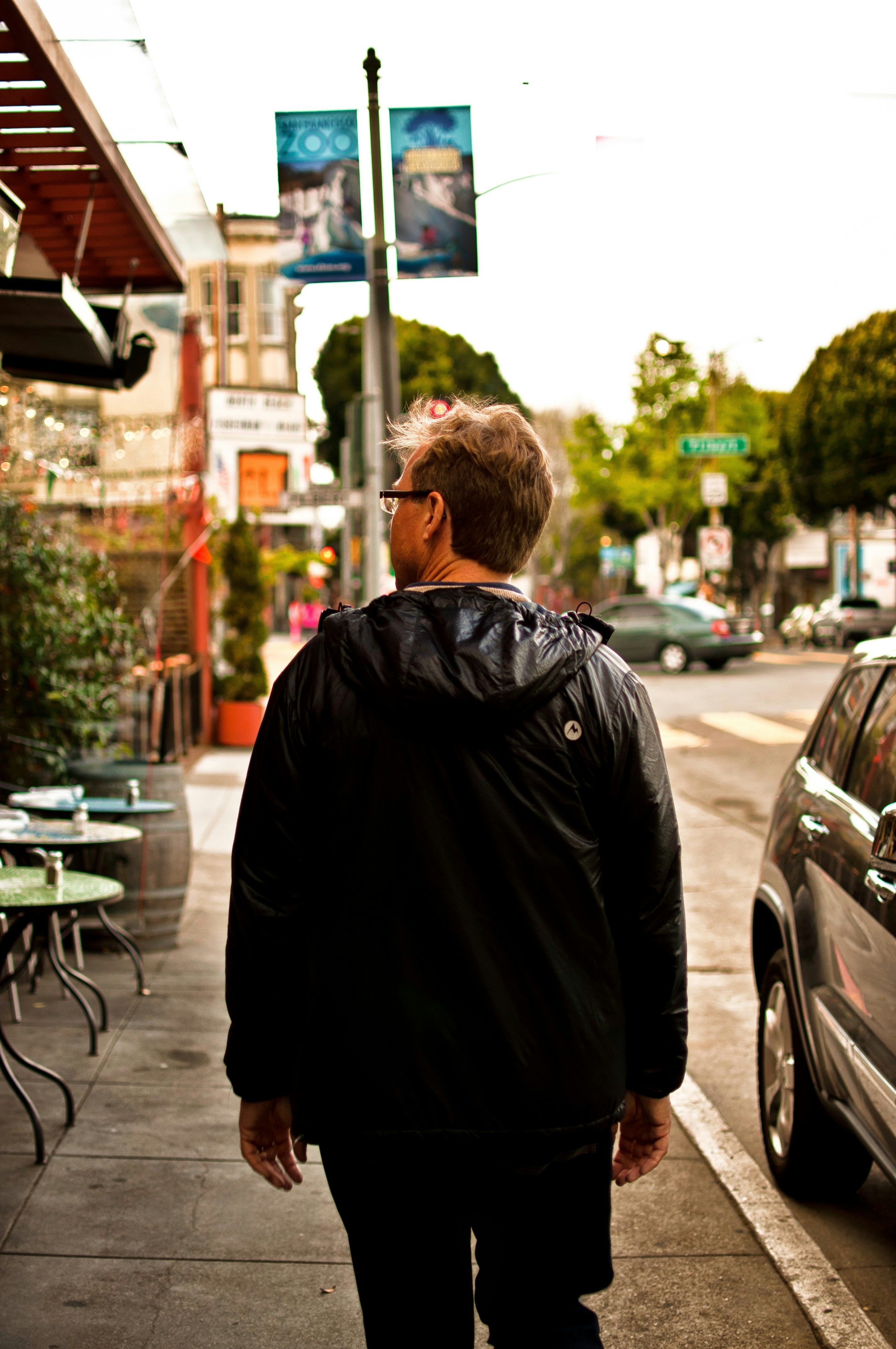 Man walking down a city street, flanked by cafes and urban greenery. The atmosphere reflects a casual urban lifestyle.