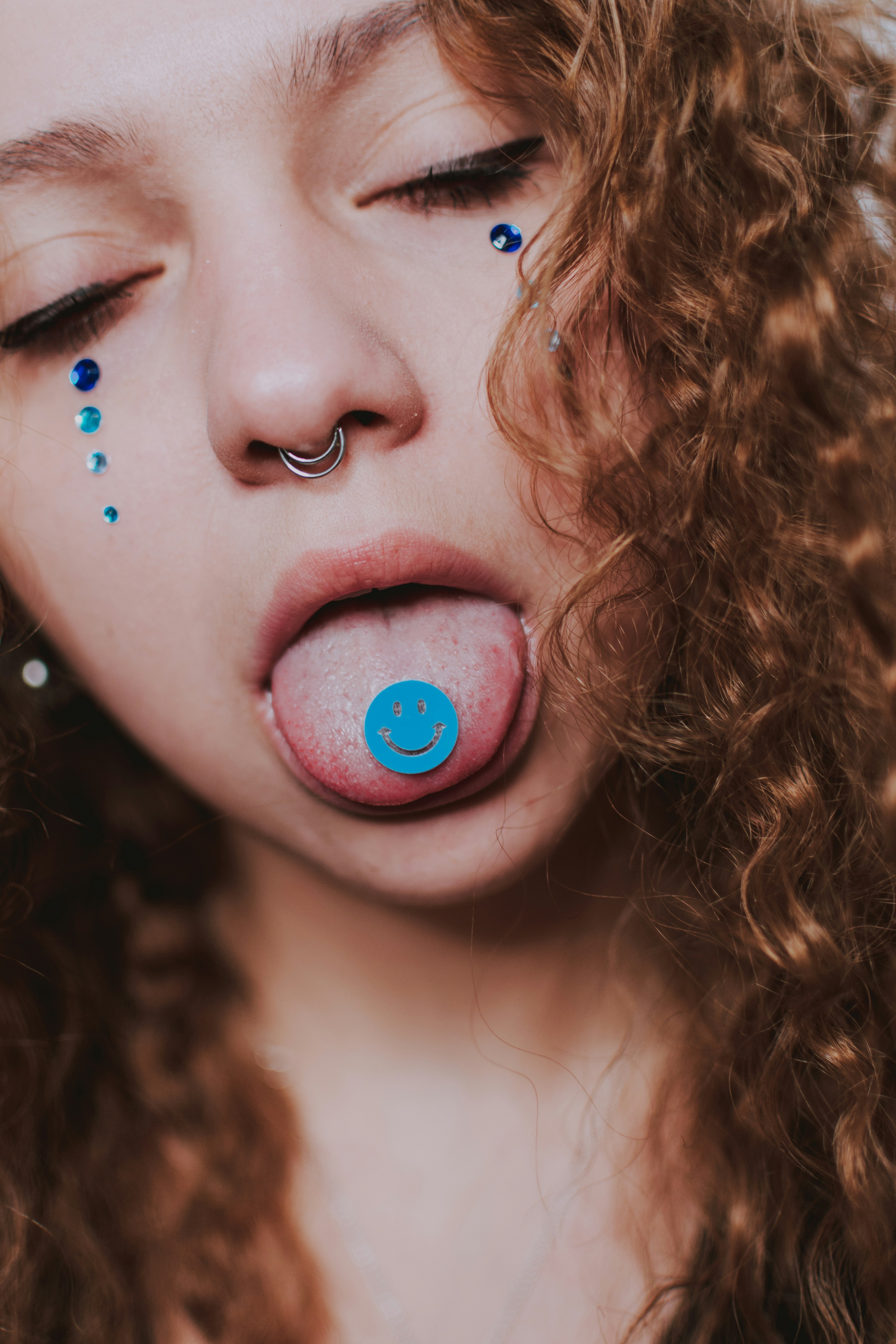 Close-up portrait showcasing a young person with a smiley face sticker on their tongue, adorned with decorative gems around their eyes.