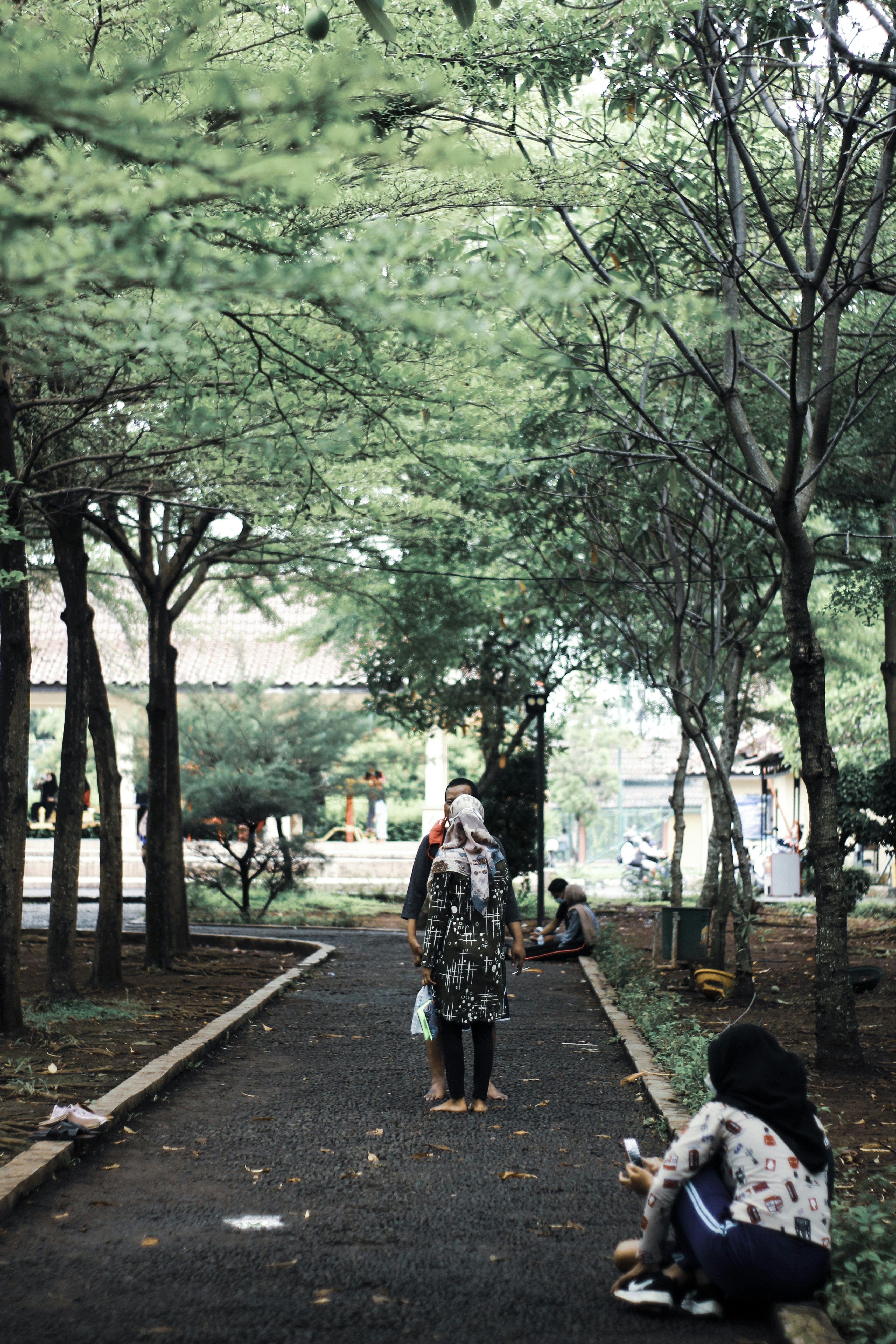 Women walking along a tree-lined path in a serene park, surrounded by lush greenery. The scene captures a moment of tranquility amidst the natural setting.
