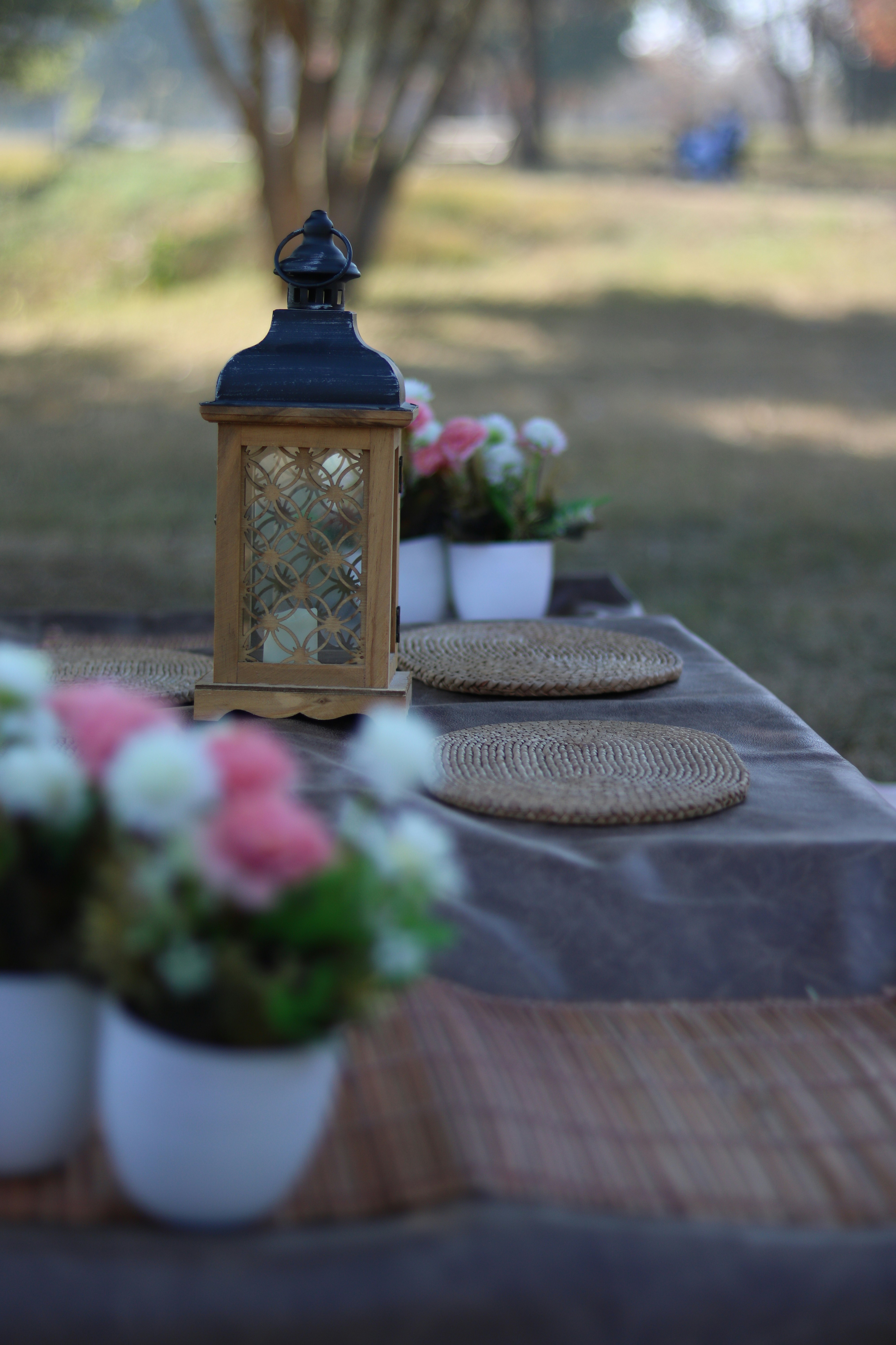 A wooden lantern stands prominently on a table adorned with woven placemats and vibrant floral arrangements in white pots. The tranquil outdoor setting enhances the inviting atmosphere.