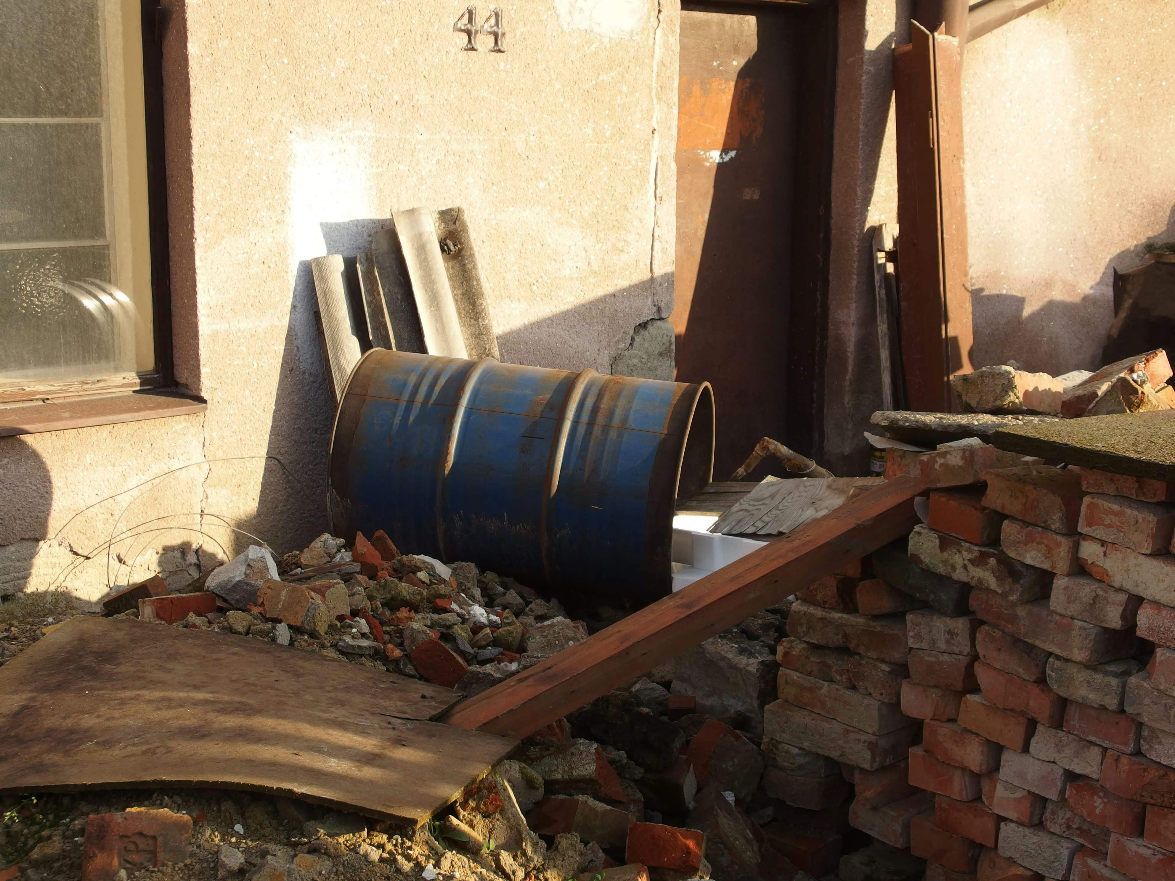 A weathered blue barrel rests amidst rubble and debris near a dilapidated building, hinting at a story of neglect and decay.