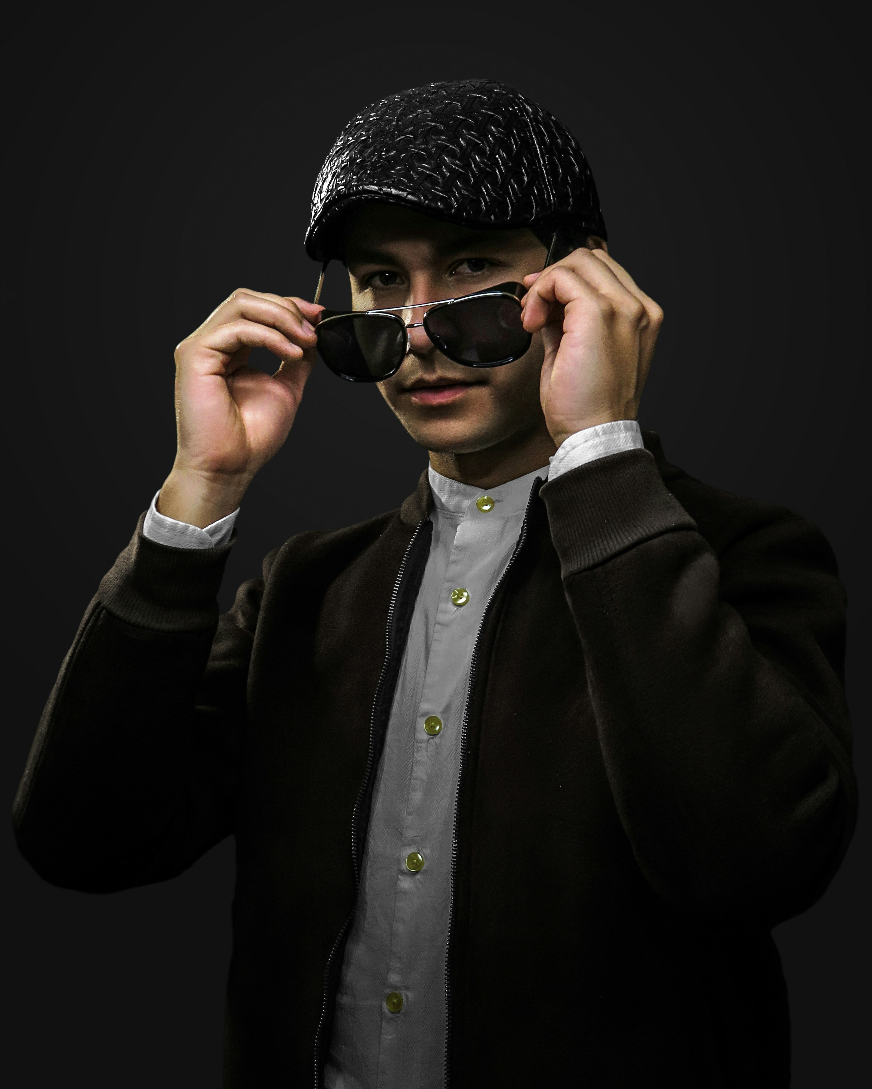 Studio portrait photograph of a young man in a dark jacket and cap, adjusting sunglasses against a black backdrop.