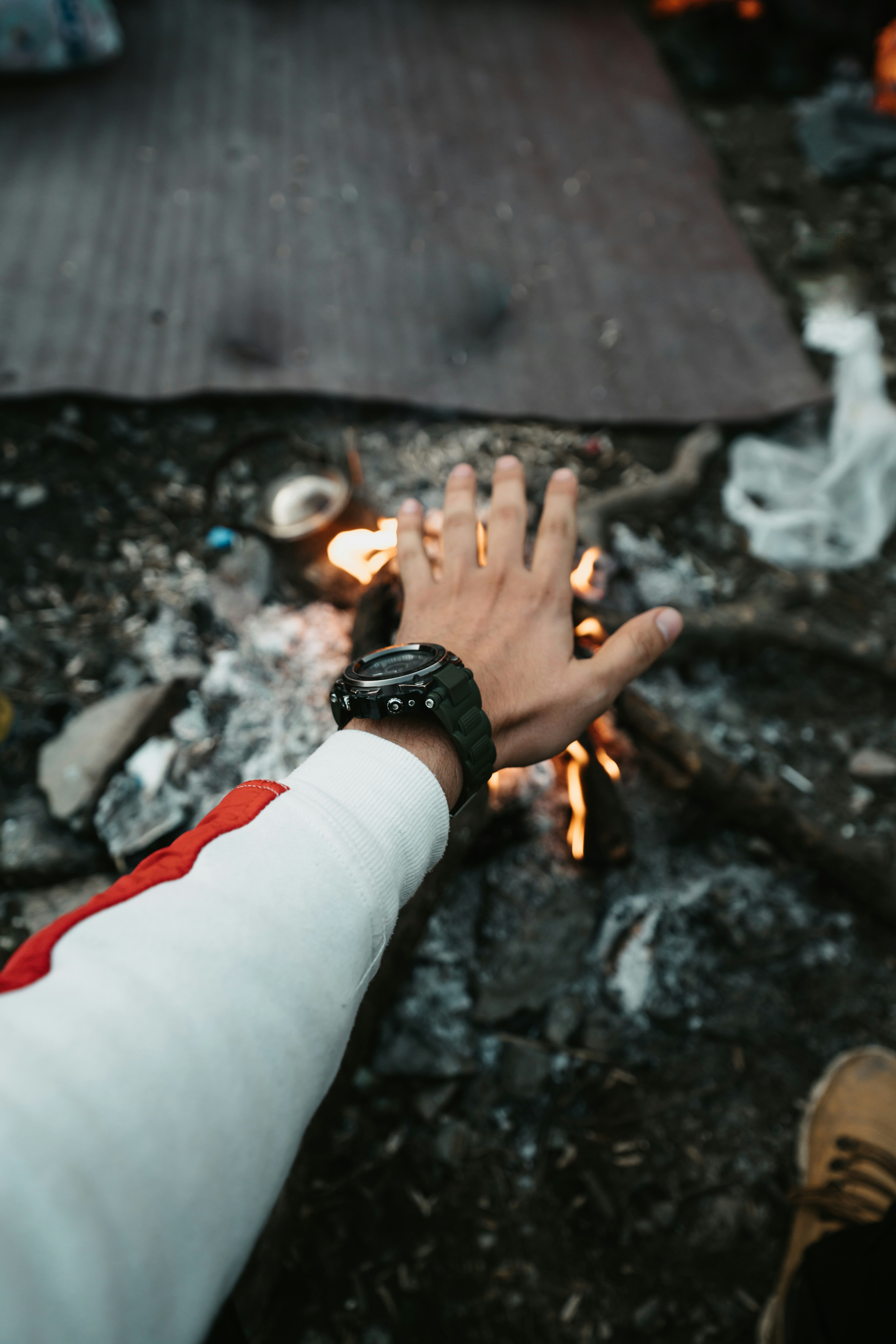 A hand reaches toward flickering flames, highlighting the warmth of a campfire amidst a rustic setting. The scene captures the essence of outdoor camaraderie.