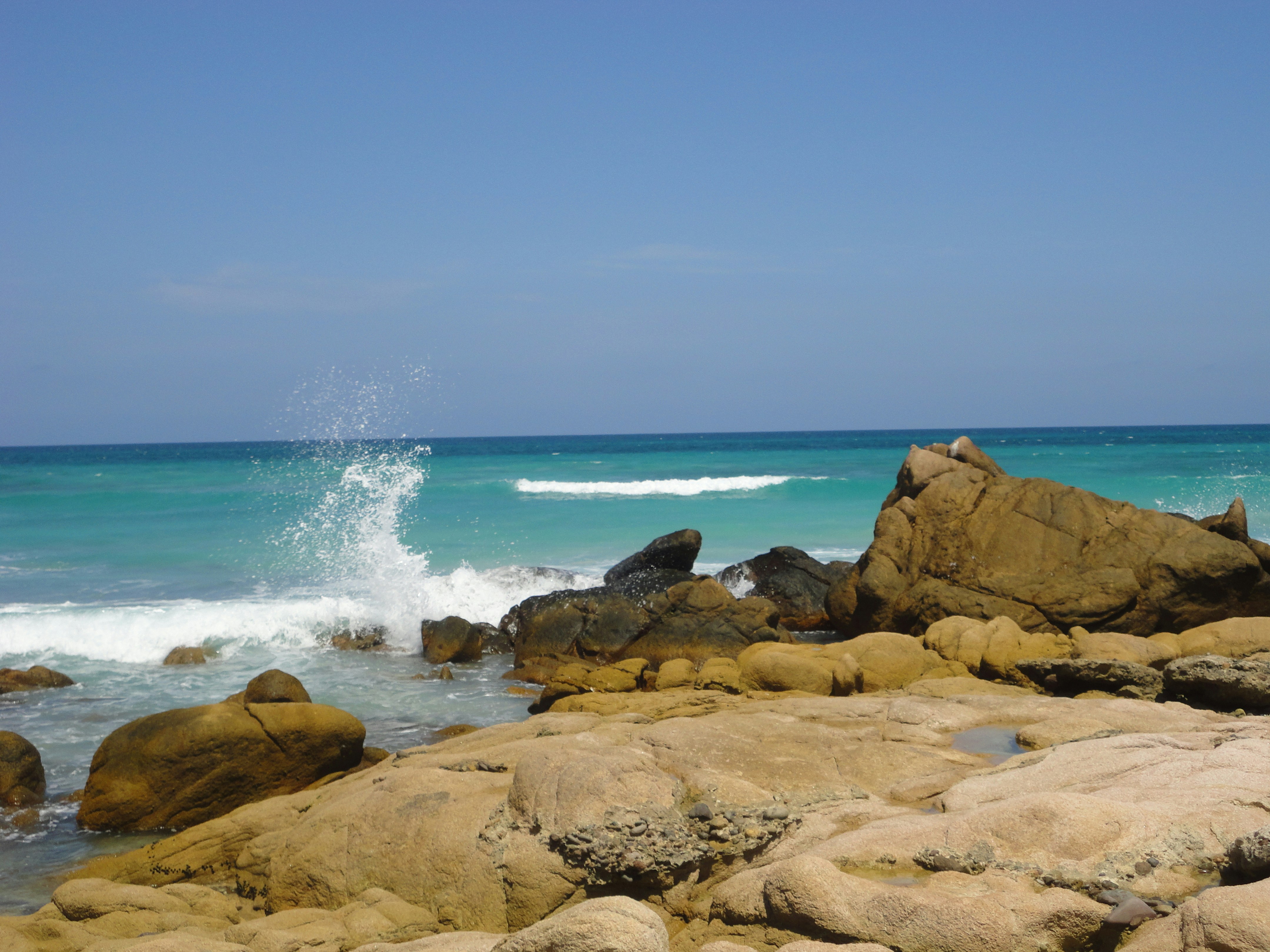 Waves splashing against large rocks on a sunny beach with a clear blue sky.