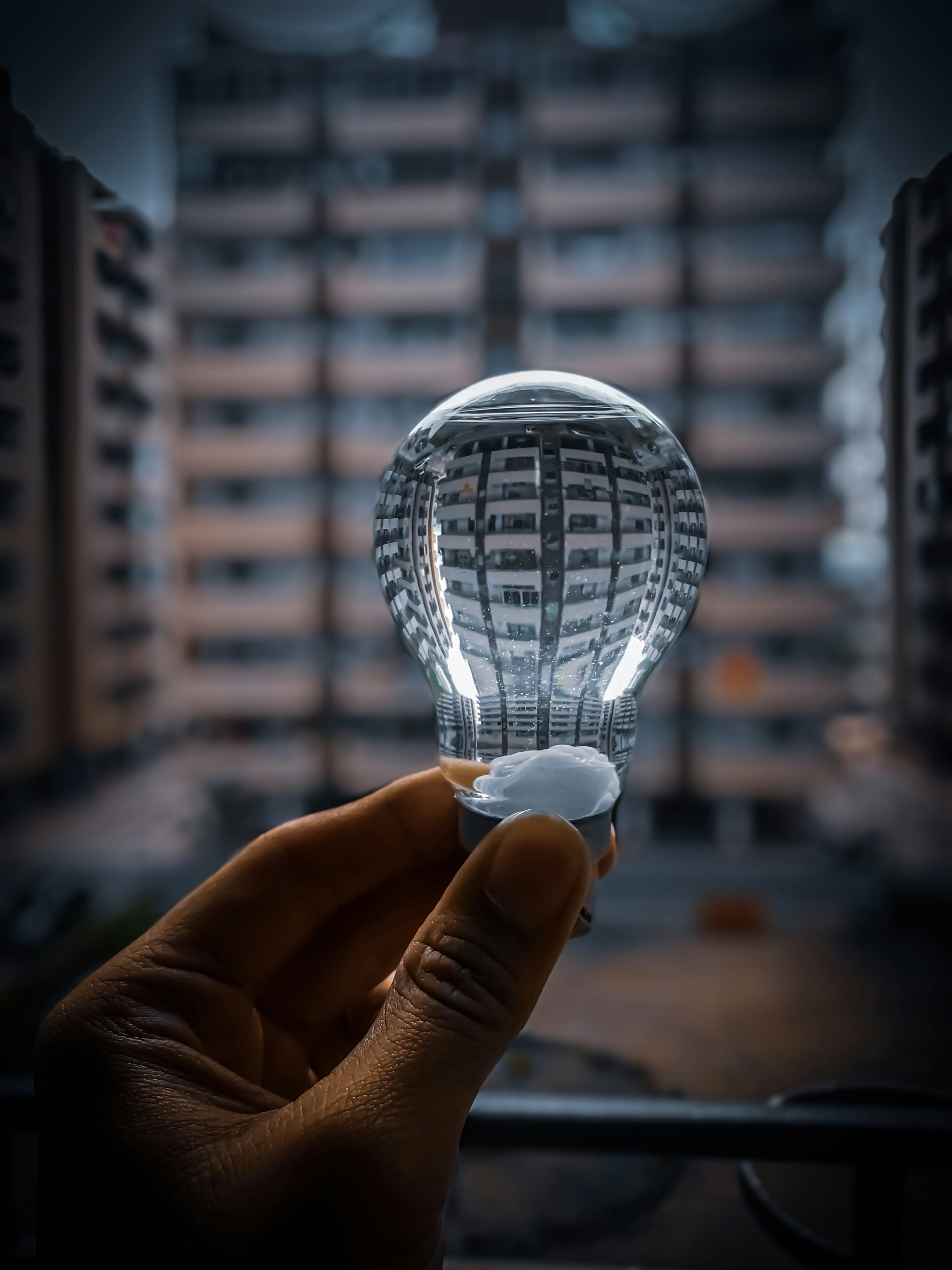 Hand holding a transparent light bulb against a backdrop of urban buildings, showcasing intricate reflections and details.