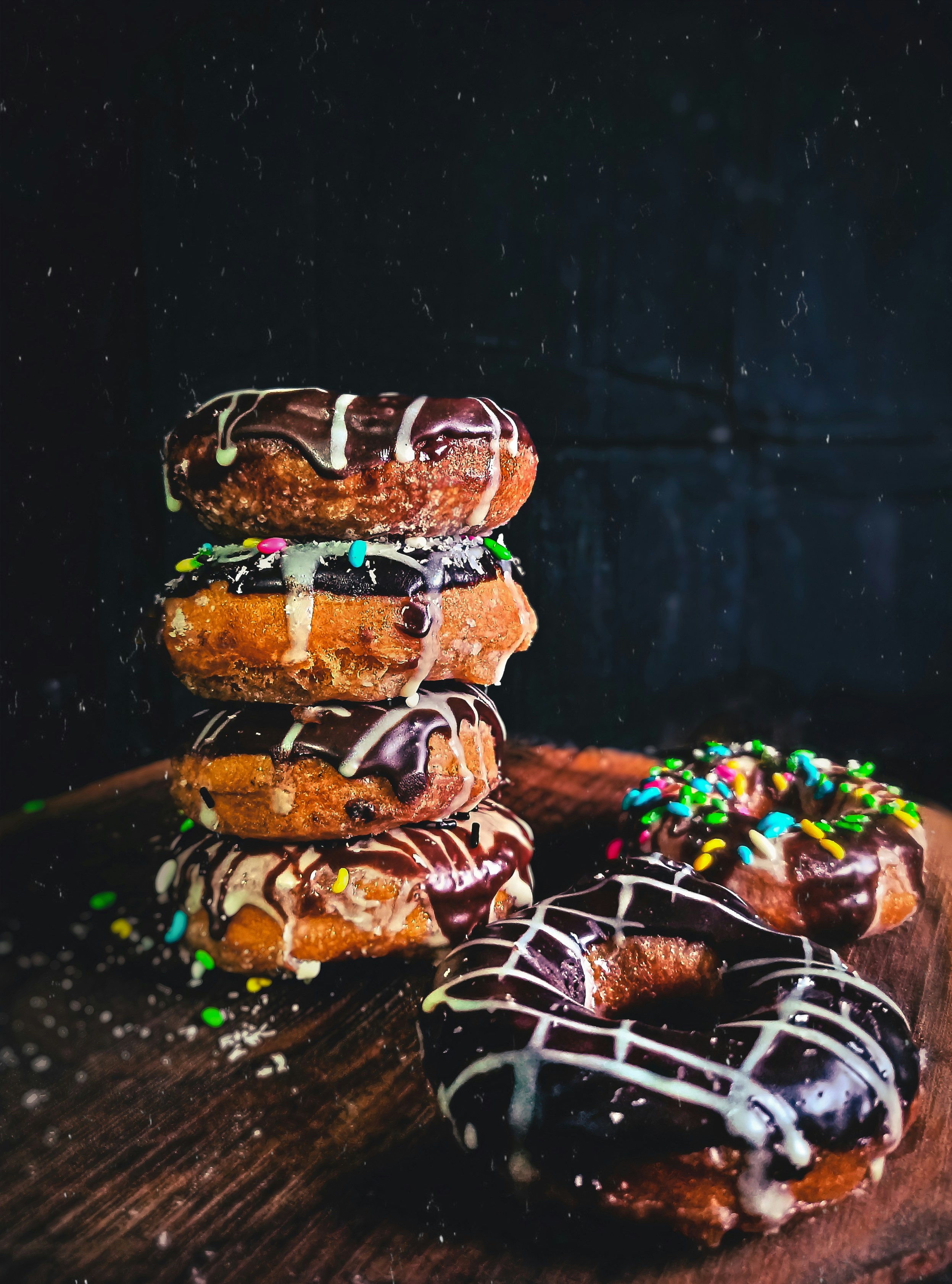 Stack of assorted doughnuts with chocolate glaze and colorful sprinkles on a wooden surface. Dark background enhances the appetizing details.