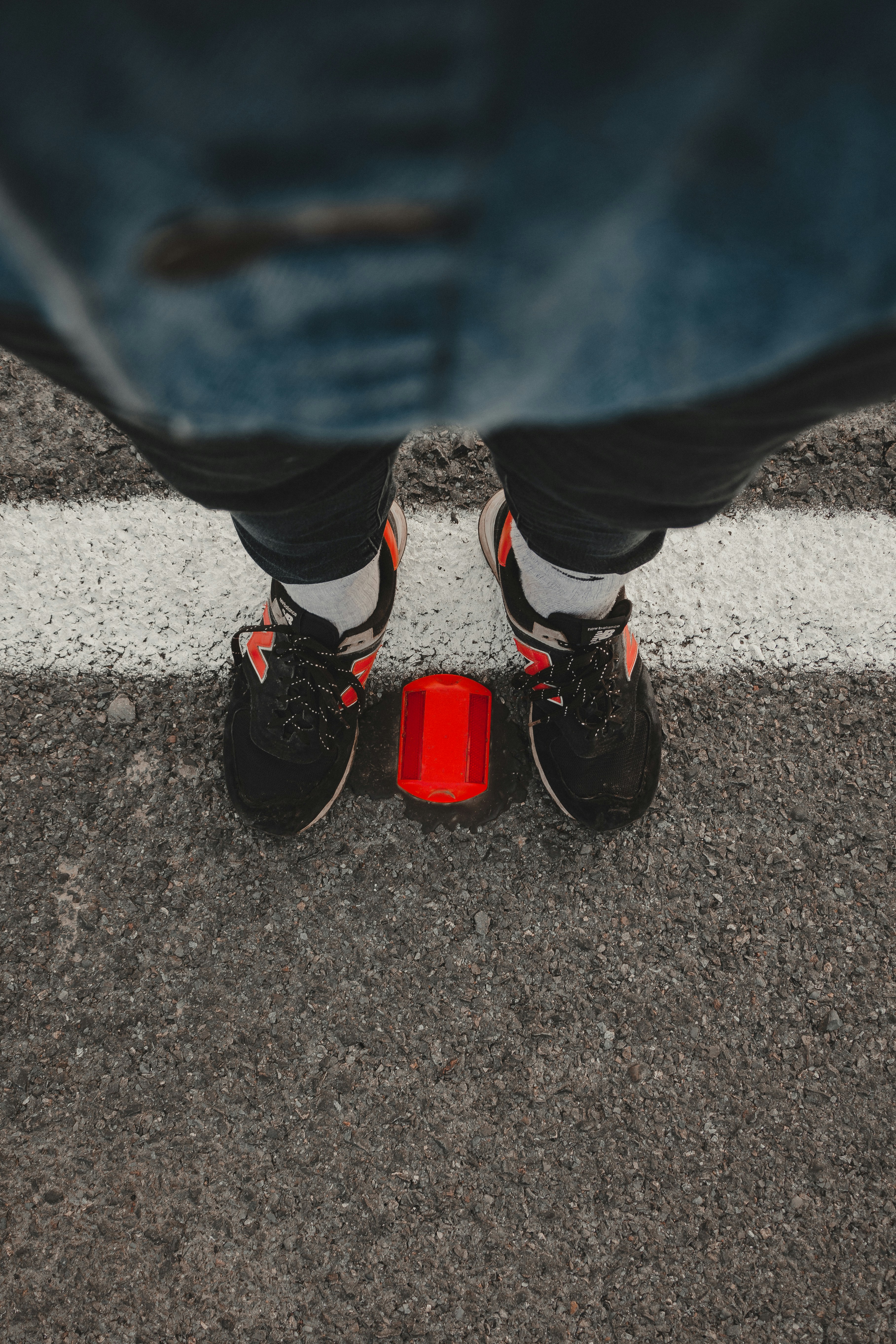 A pair of black sneakers stands on a gravel road next to a small red object, framed by a white line. The perspective emphasizes the footwear and the unusual item.