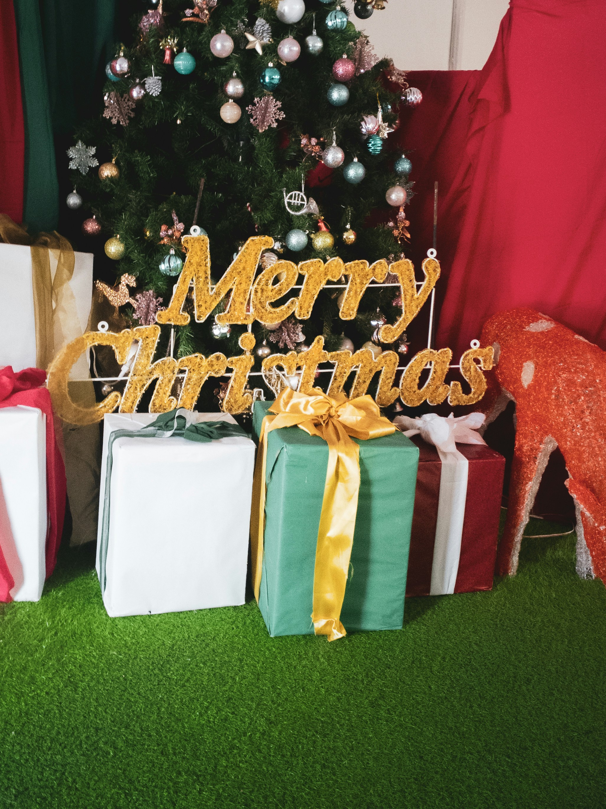 Colorful presents sit under a decorated Christmas tree with a glittering 'Merry Christmas' sign in warm lighting.