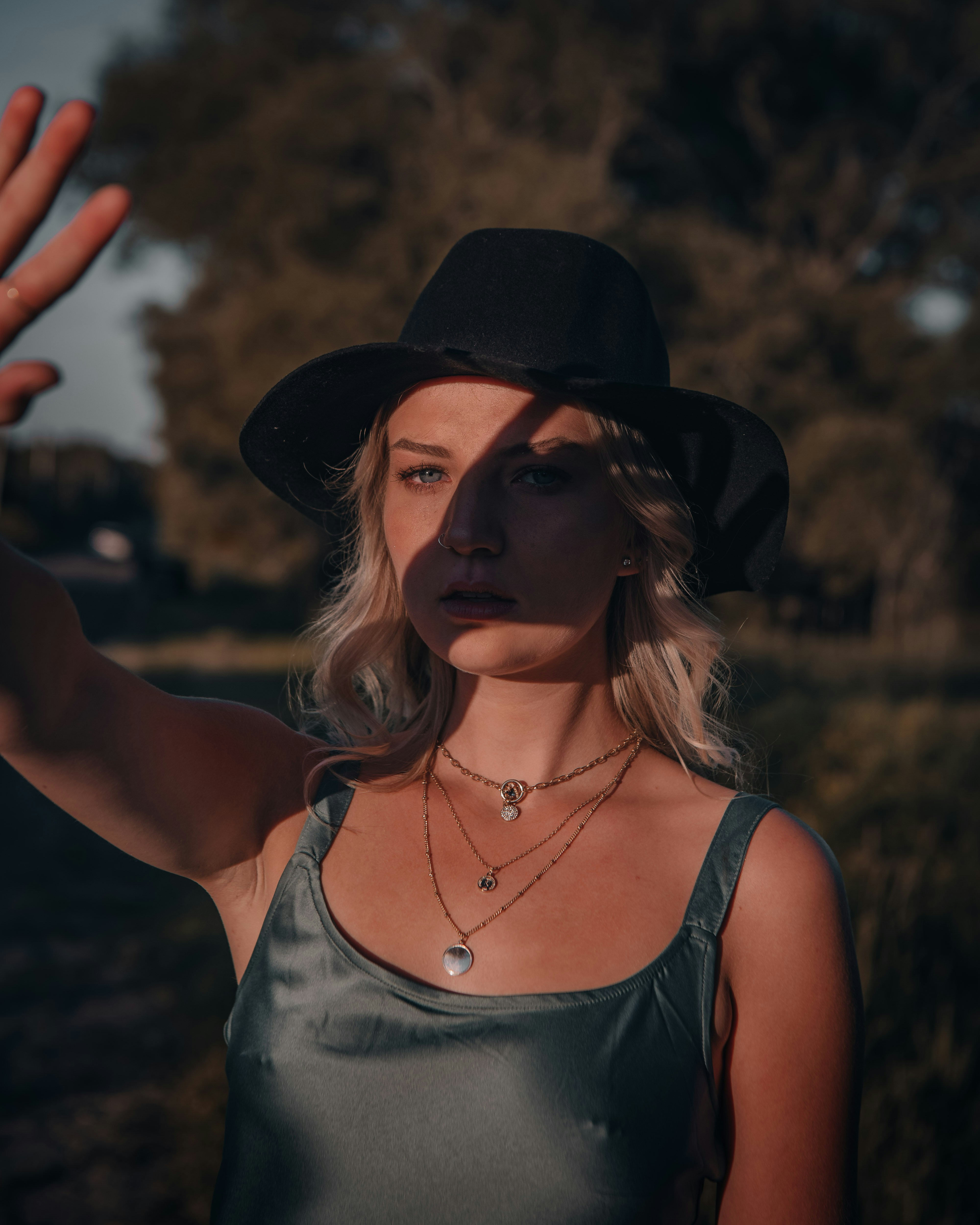 Portrait photograph of a woman in a satin top wearing a wide-brim hat, her face half-lit and a hand raised against a blurred natural backdrop.