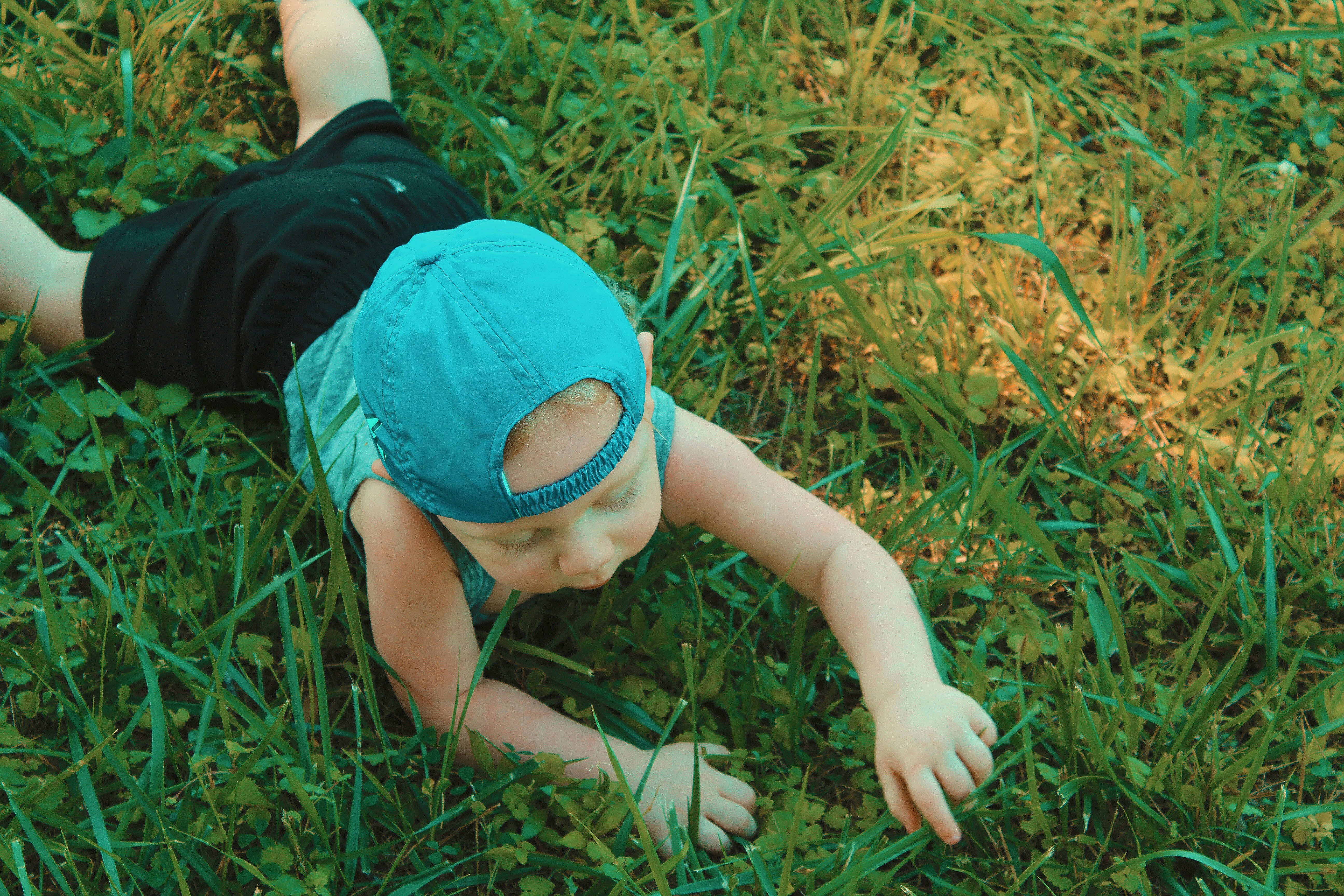 Toddler in a blue cap crawling through lush green grass, engaging with the natural environment.