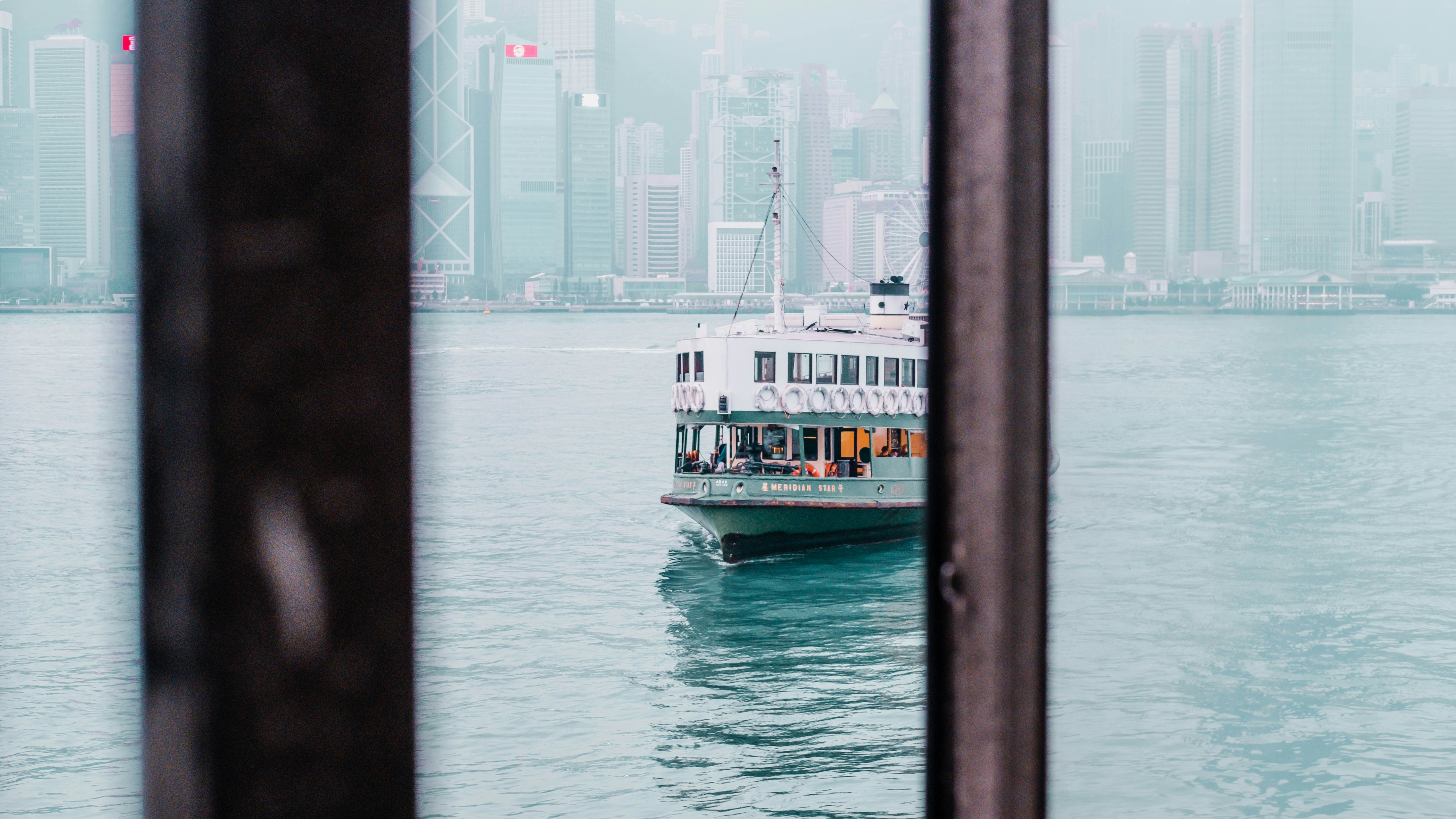 Ferry gliding through calm waters framed by metallic structures, with a hazy city skyline in the background.