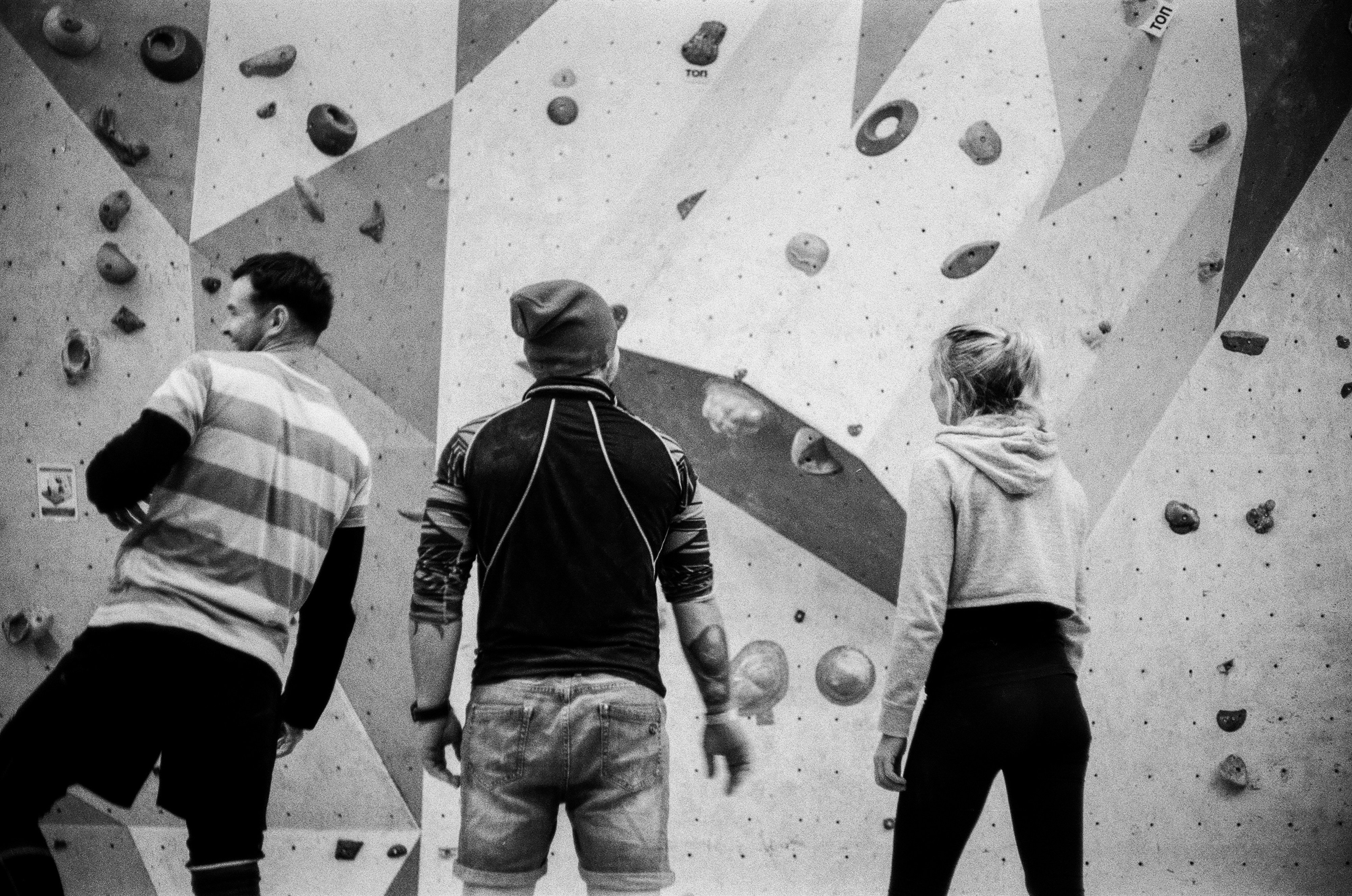 Three climbers stand with backs to the camera before a faceted climbing wall, chalk on their hands. This black-and-white photograph captures a quiet moment before the ascent.