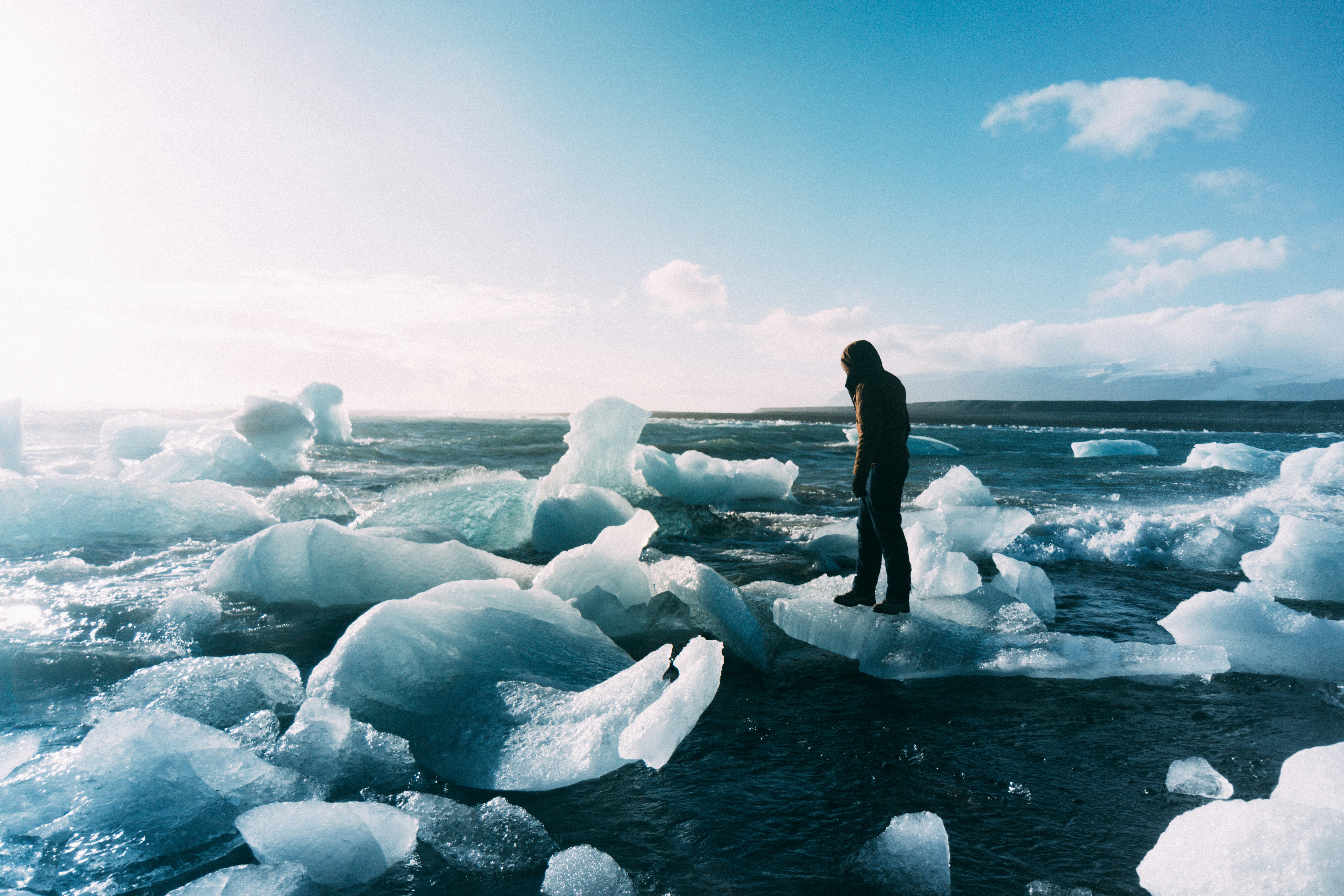 A figure stands amidst floating icebergs, gazing across a serene, glistening ocean under a bright sky.