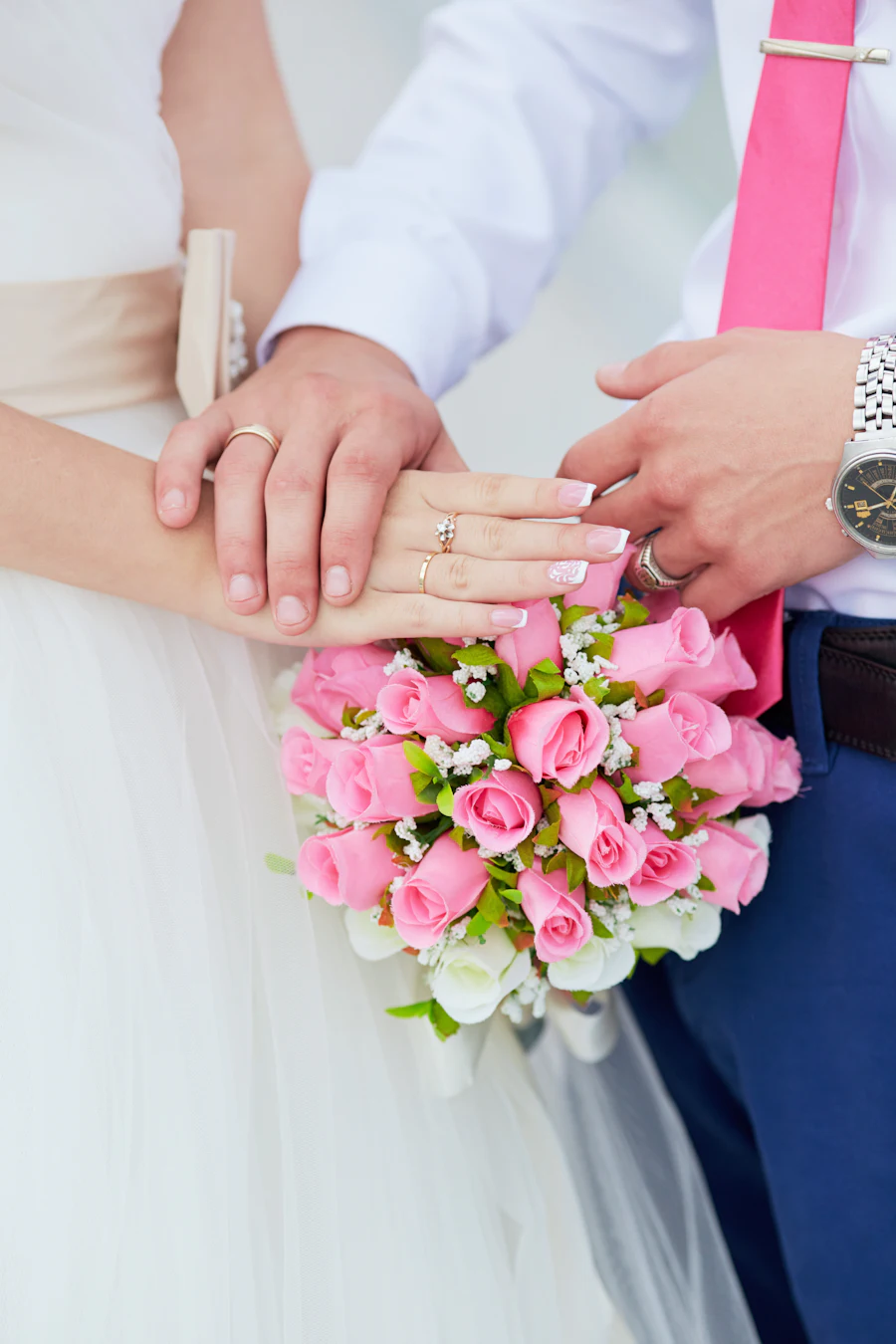 Bride and groom’s hands stacked together above a bouquet of pink and white roses, with wedding attire and bright pink tie visible. Close-up of a romantic ceremony detail.