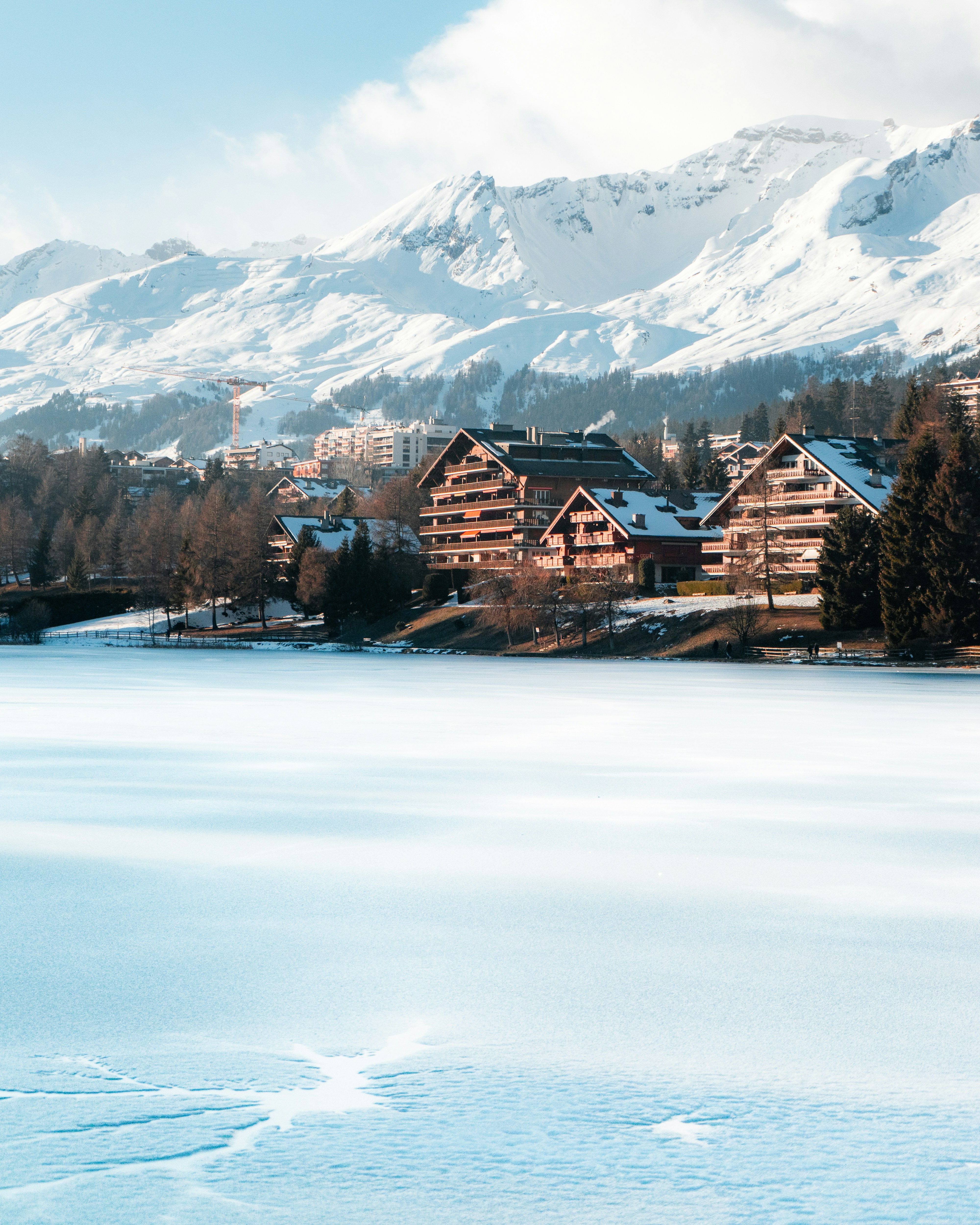 Charming wooden chalets nestled along a frozen lake, framed by majestic snow-covered mountains under a clear blue sky.