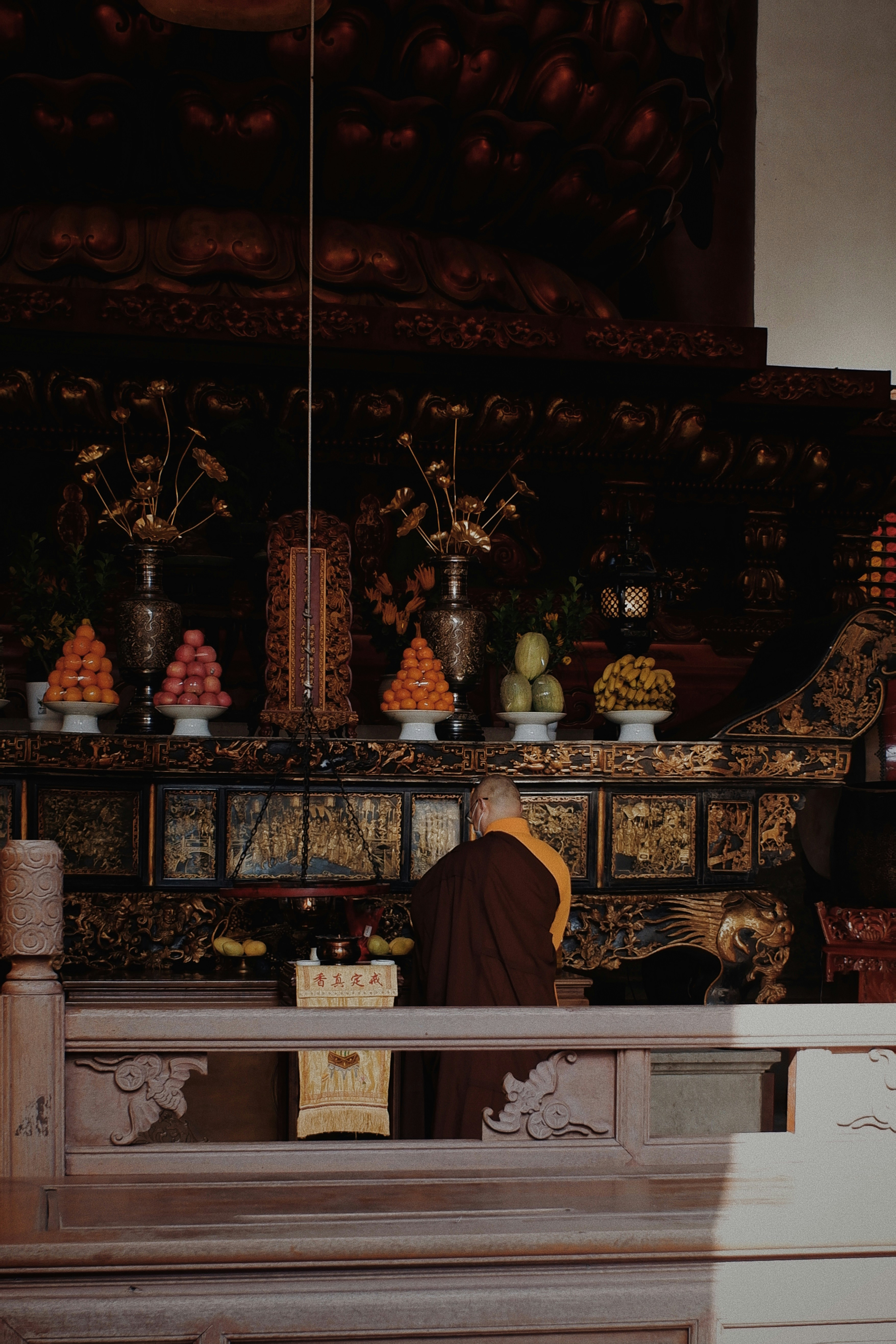 Monk in a brown robe stands with back to the camera before an ornate altar lined with stacked fruit offerings and gilded carvings.