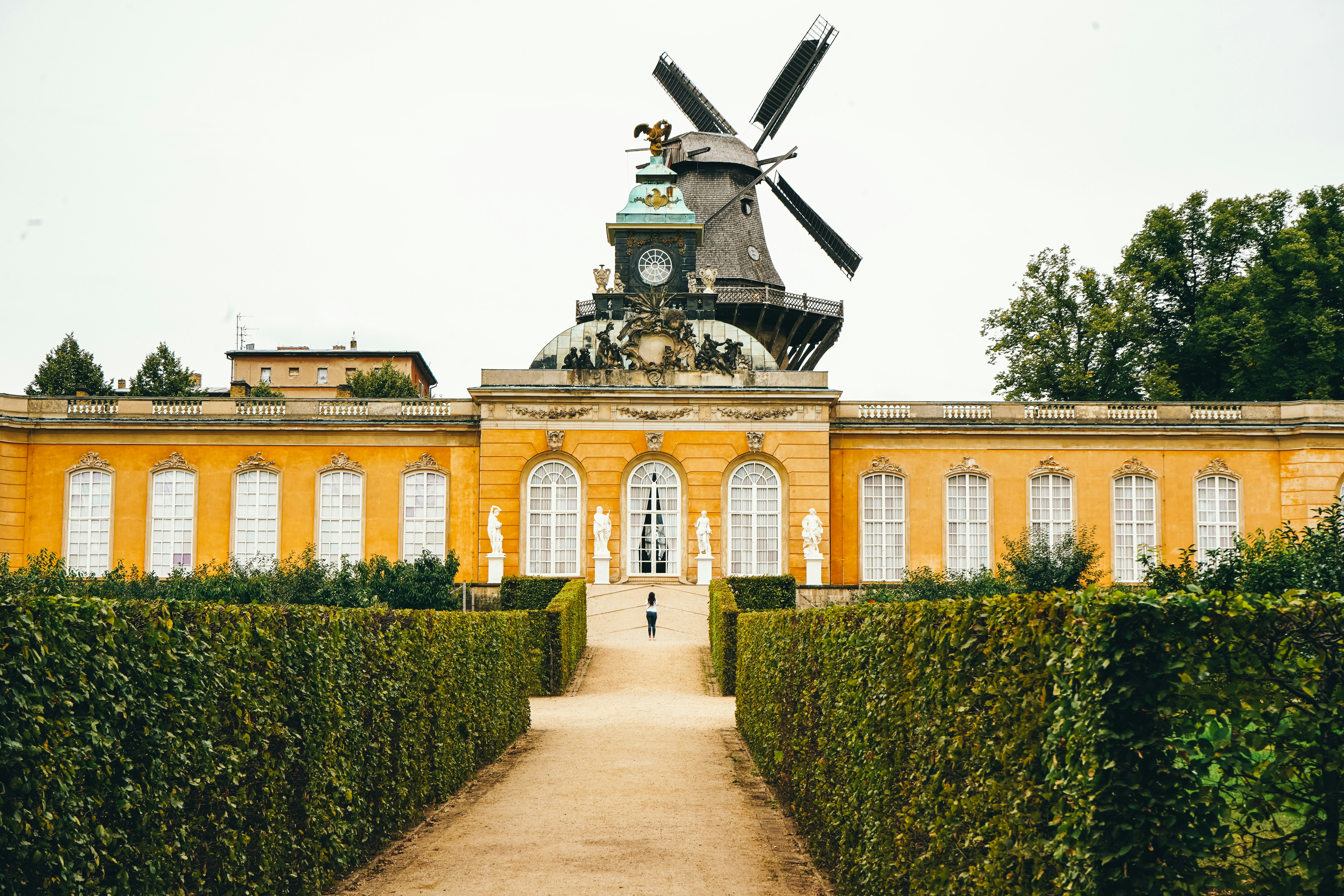 Historic windmill perched atop a grand yellow building with manicured hedges lining a sandy path.
