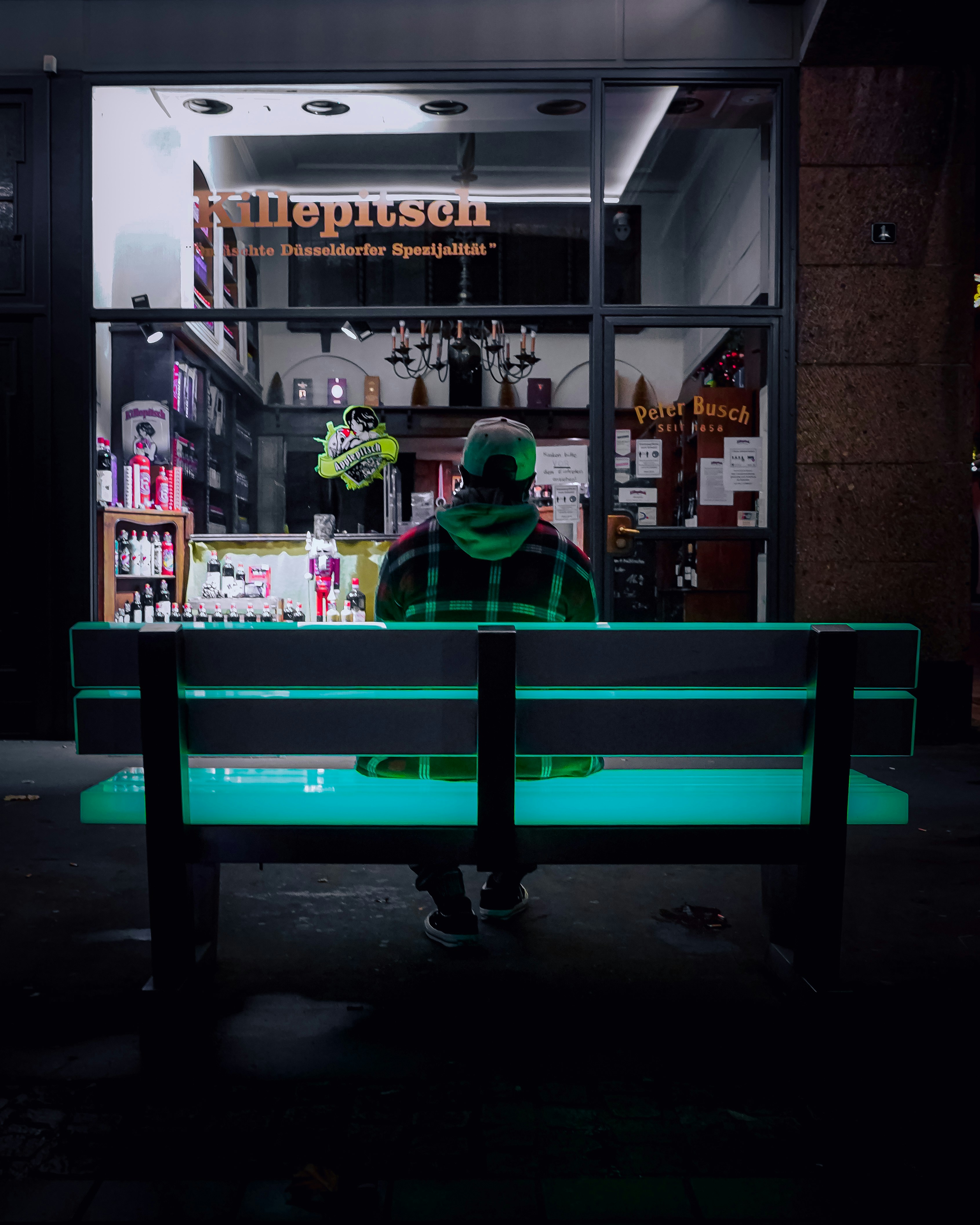 A person sits on a glowing bench, gazing at a storefront filled with colorful products, evoking a sense of contemplation in an urban setting.
