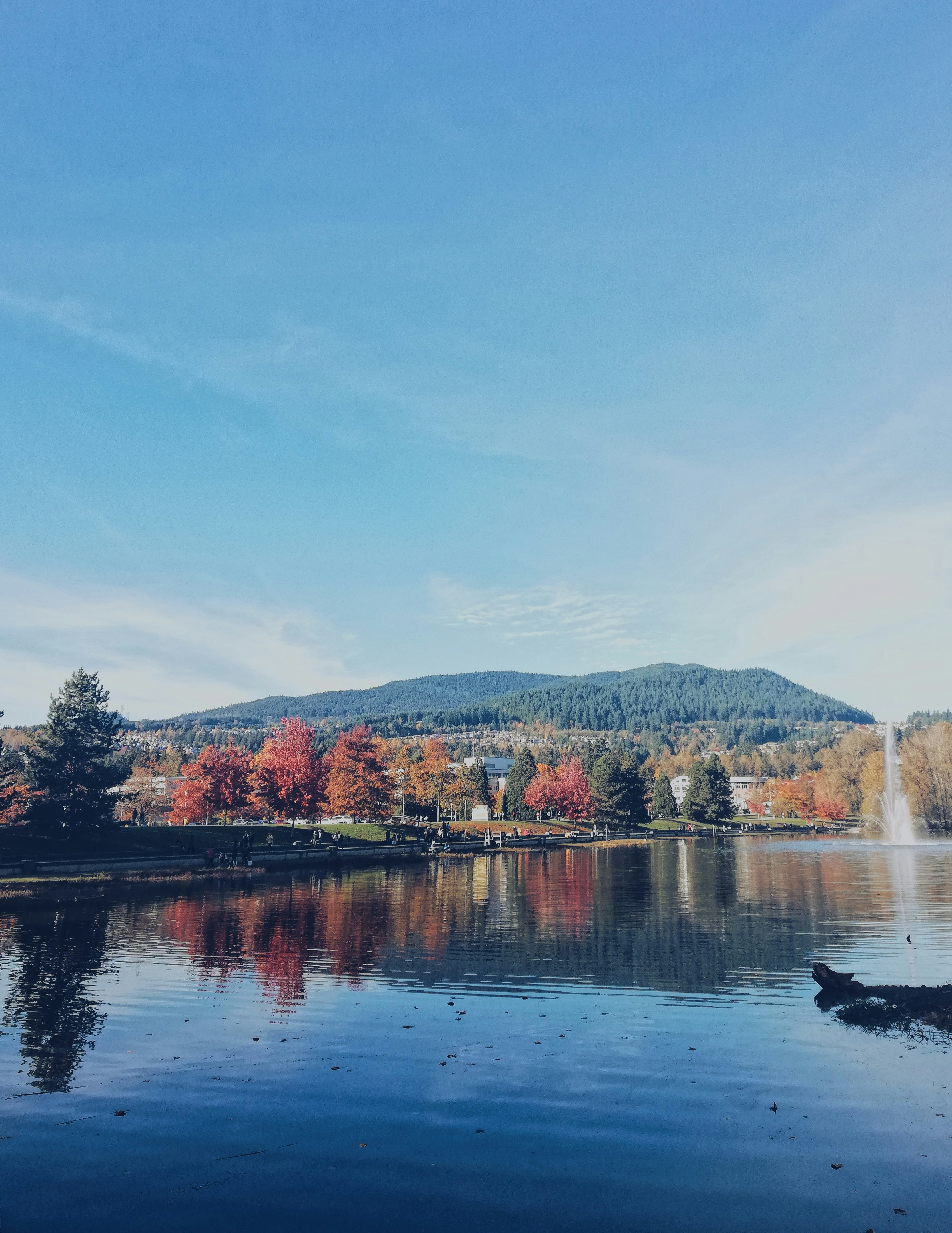 Calm lakeside scene in autumn, with colorful trees along the shore reflected in the still water and distant hills under a blue sky.