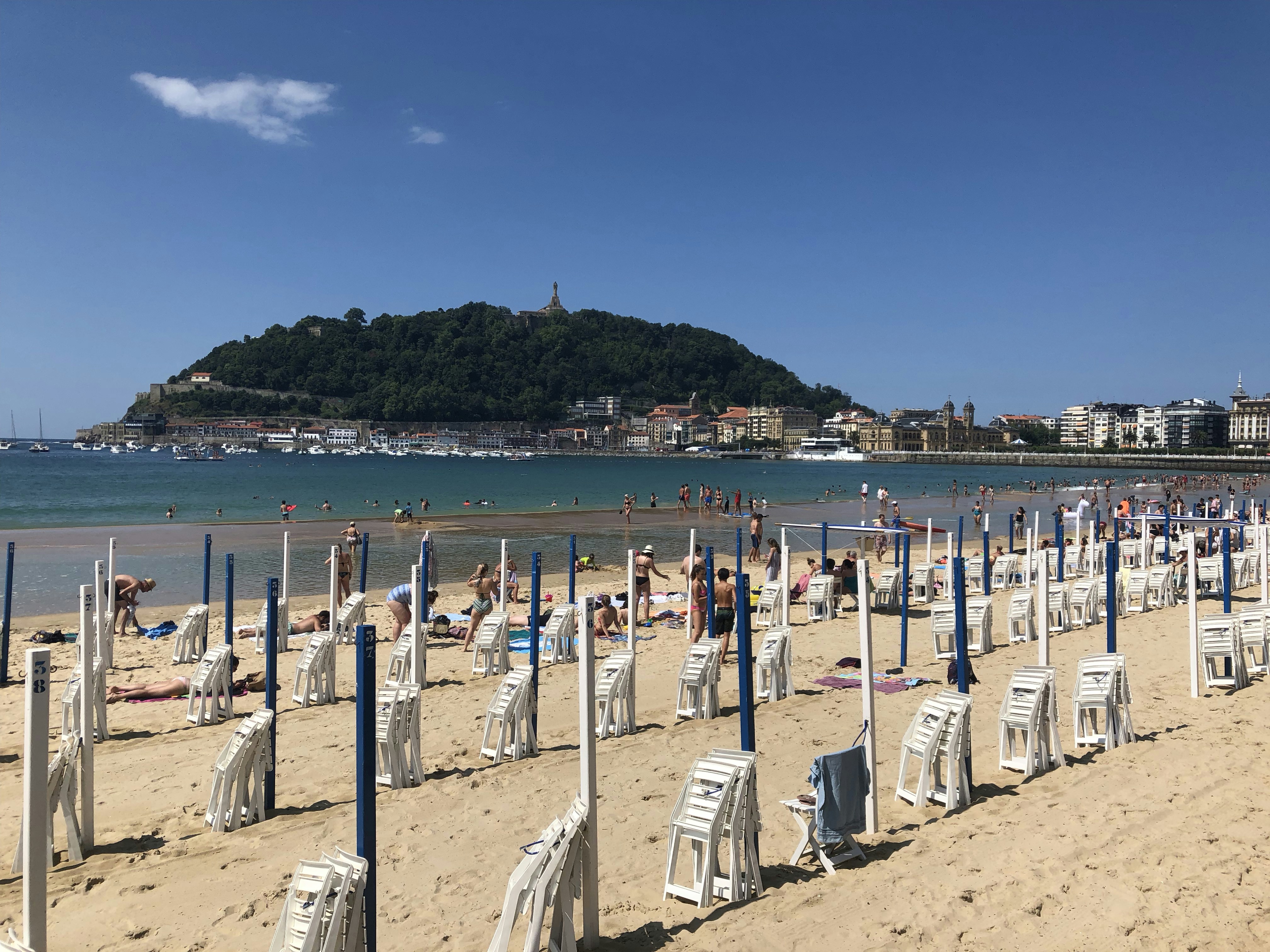 Rows of empty beach chairs on a sunny sandy shore with a verdant hill and coastal cityscape in the background.