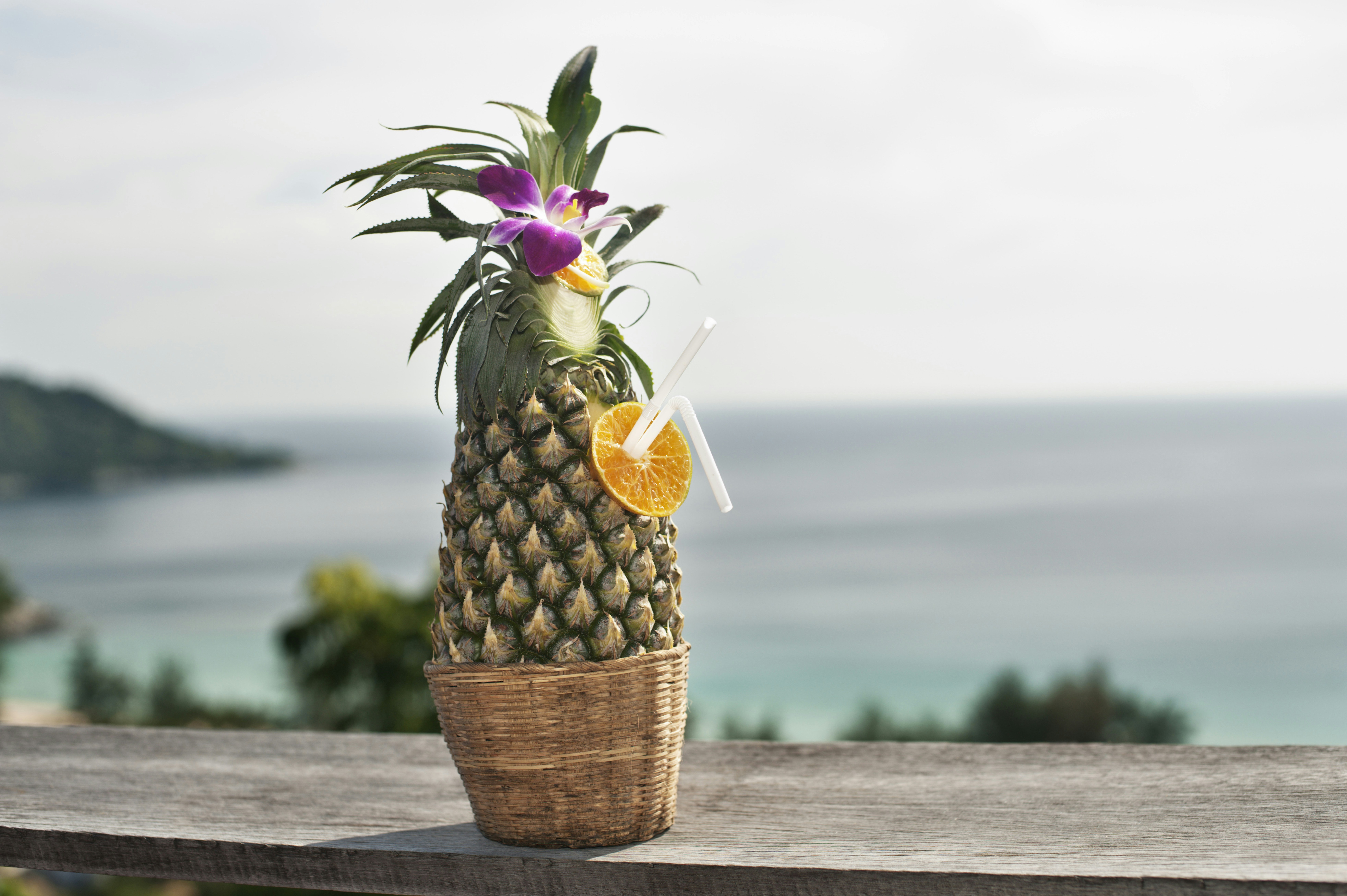 A pineapple cocktail garnished with flowers and citrus sits on a wooden railing, overlooking a serene ocean view.
