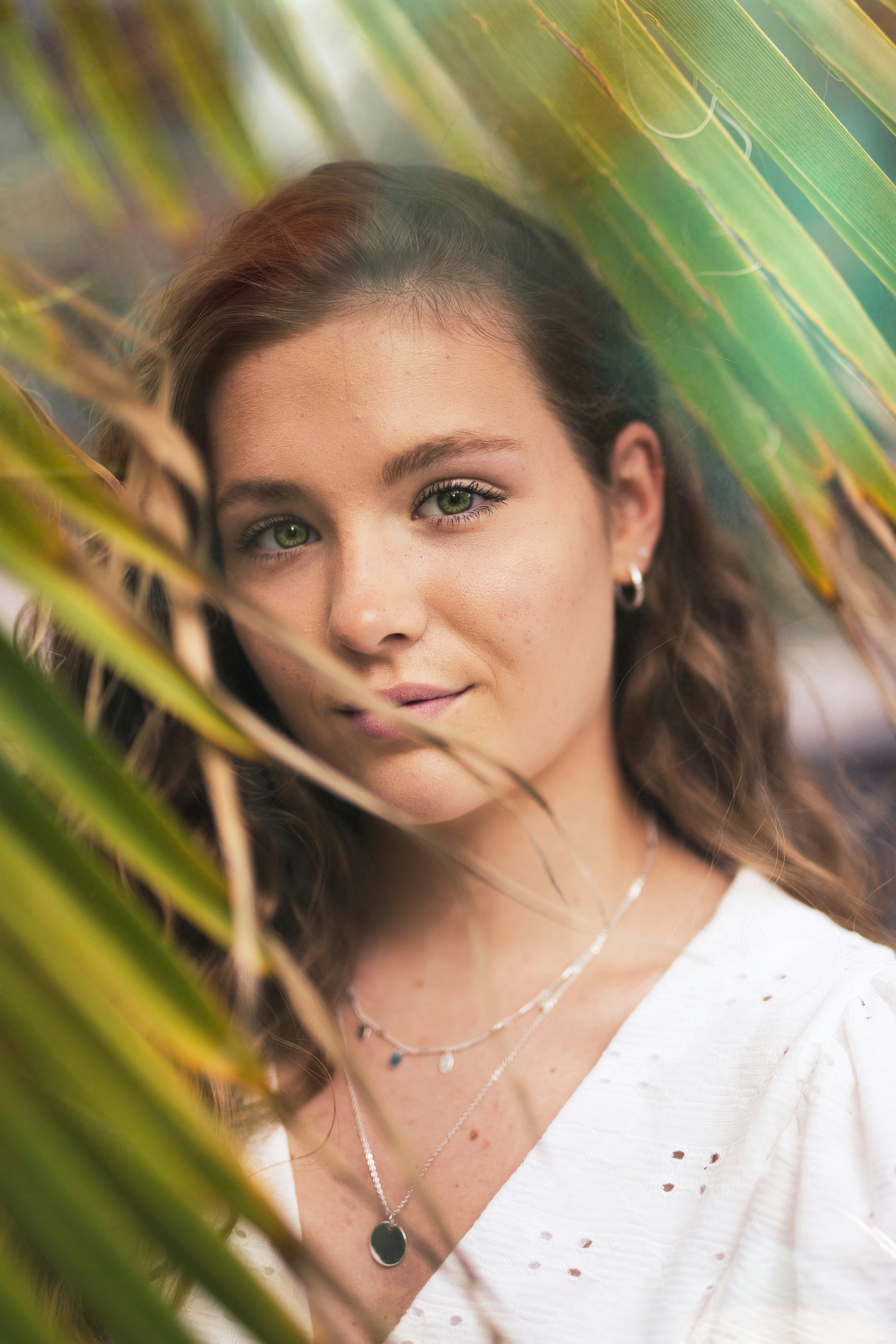 A young woman gazes softly through vibrant green palm fronds, showcasing her natural beauty and serene expression.