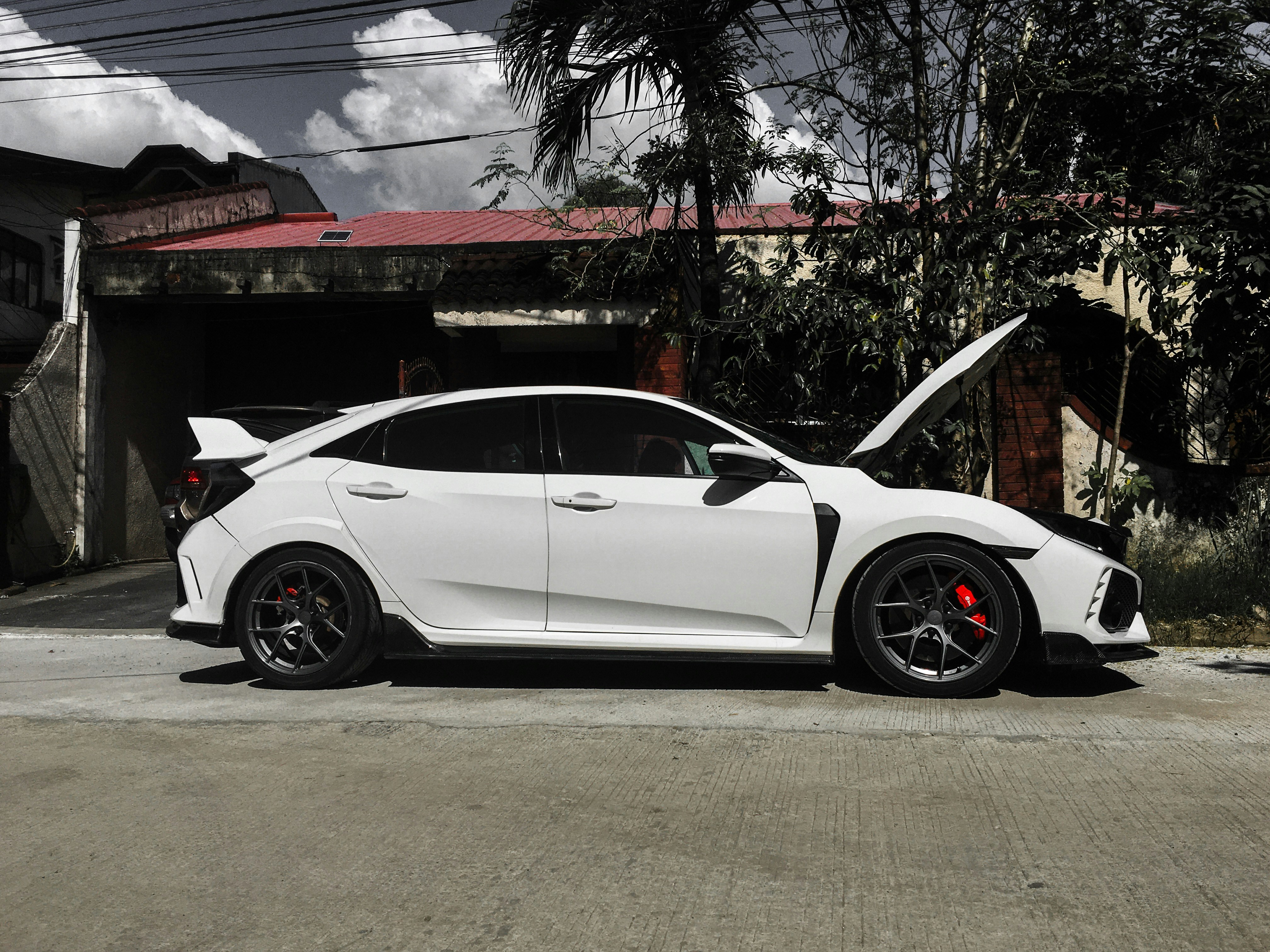 Modified white hatchback with an open hood, showcasing its engine. The car is parked on a street with a backdrop of greenery and urban elements.