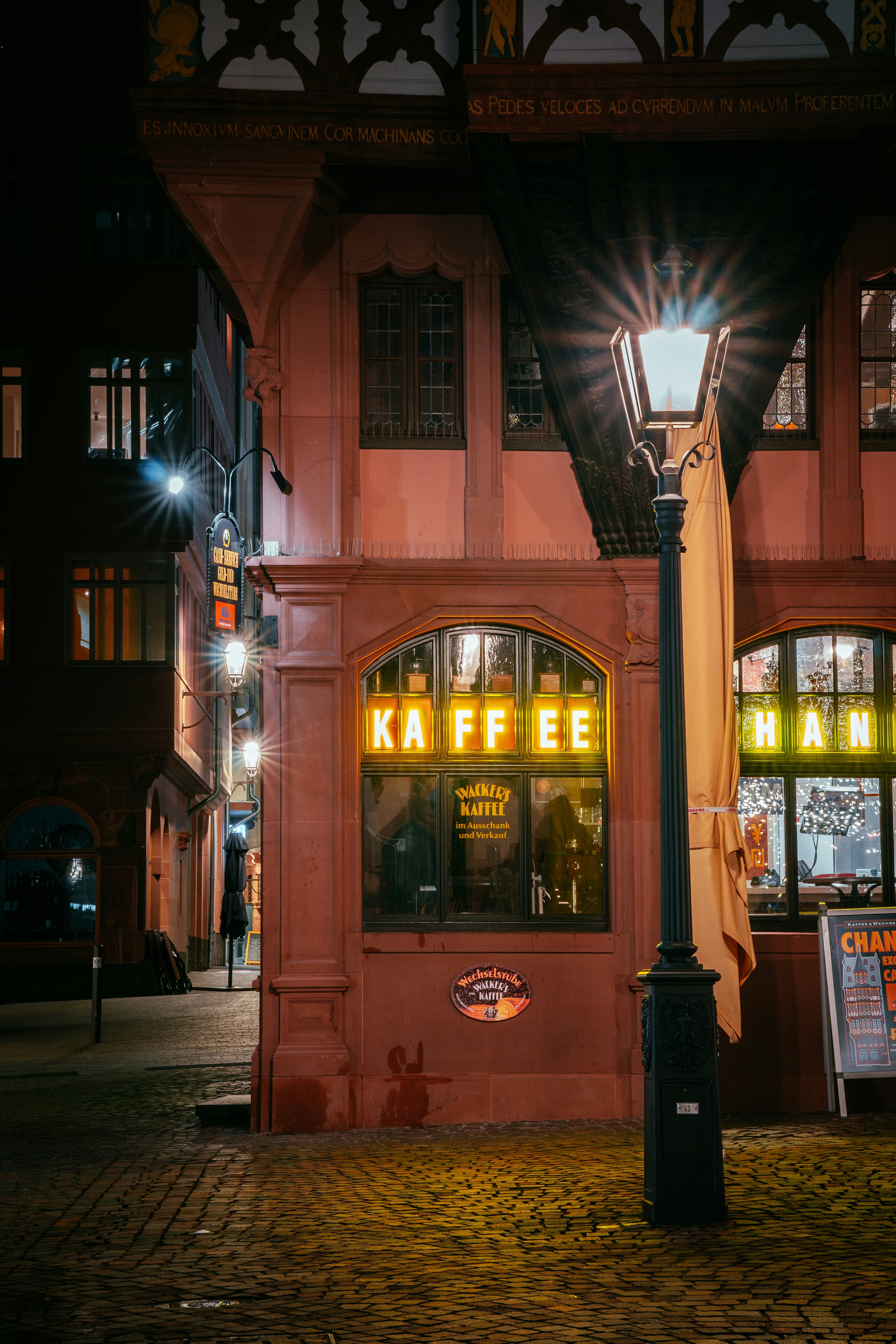 A cozy café exterior with glowing neon signs reading 'KAFFEE' and 'Waffeln' under warm streetlights, creating an inviting atmosphere in a cobblestone square.