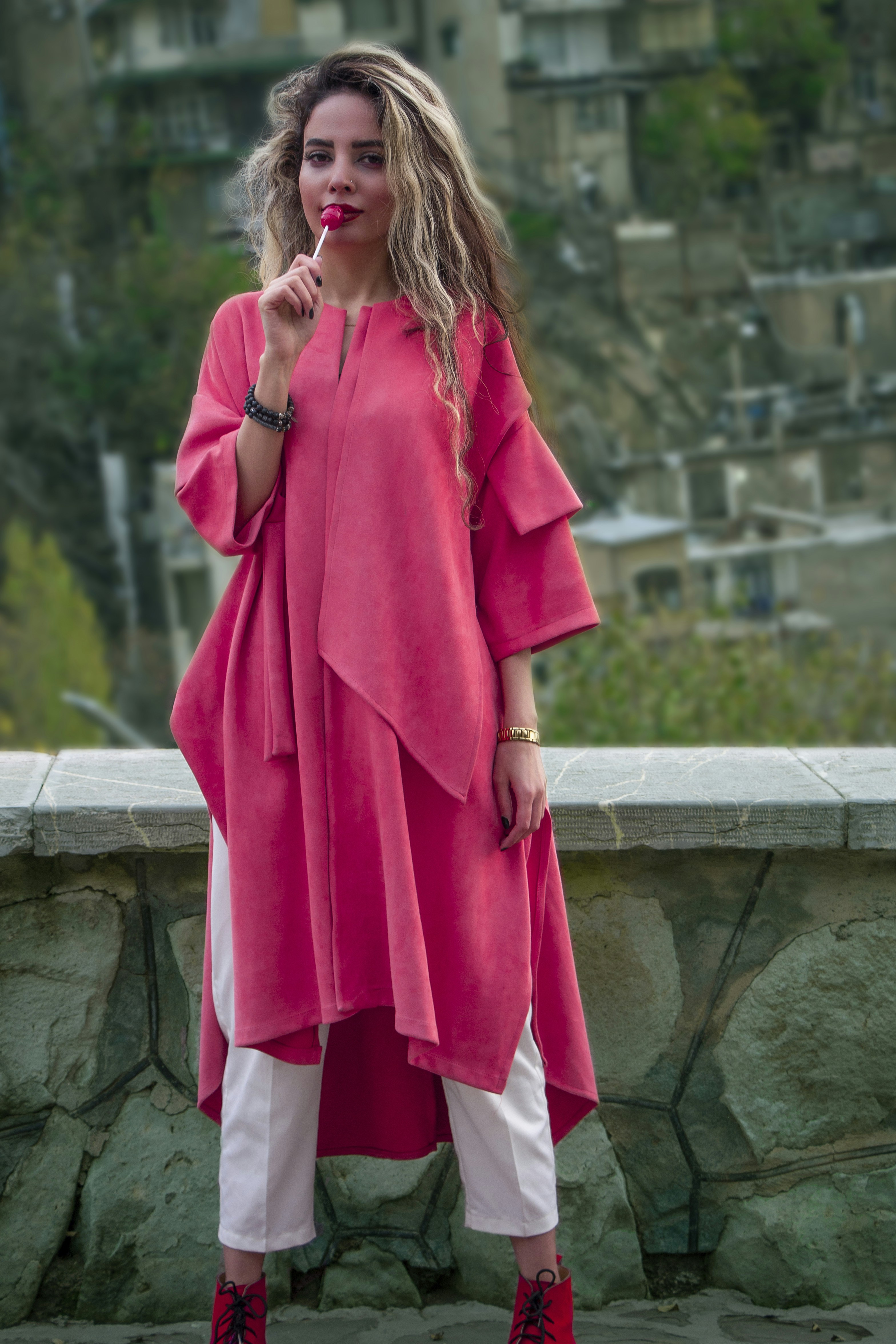 A woman in a flowing pink garment stands pensively, holding a lollipop against a blurred urban backdrop. Her stylish outfit contrasts with the muted background, adding a modern flair.