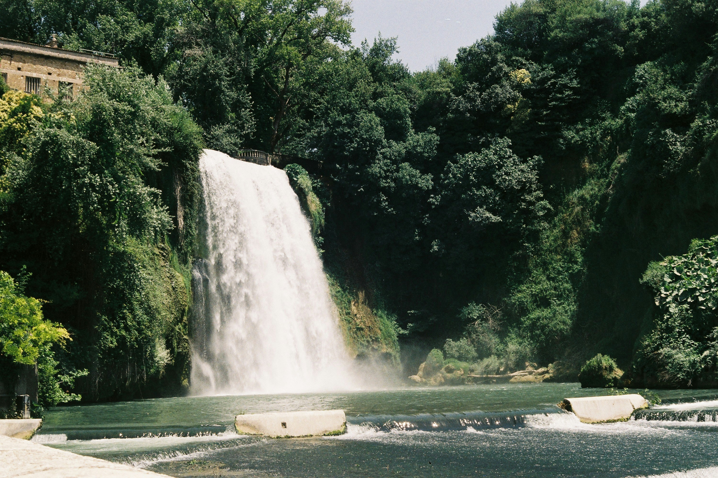 Waterfall flowing into a serene pool surrounded by dense green trees under a clear sky.