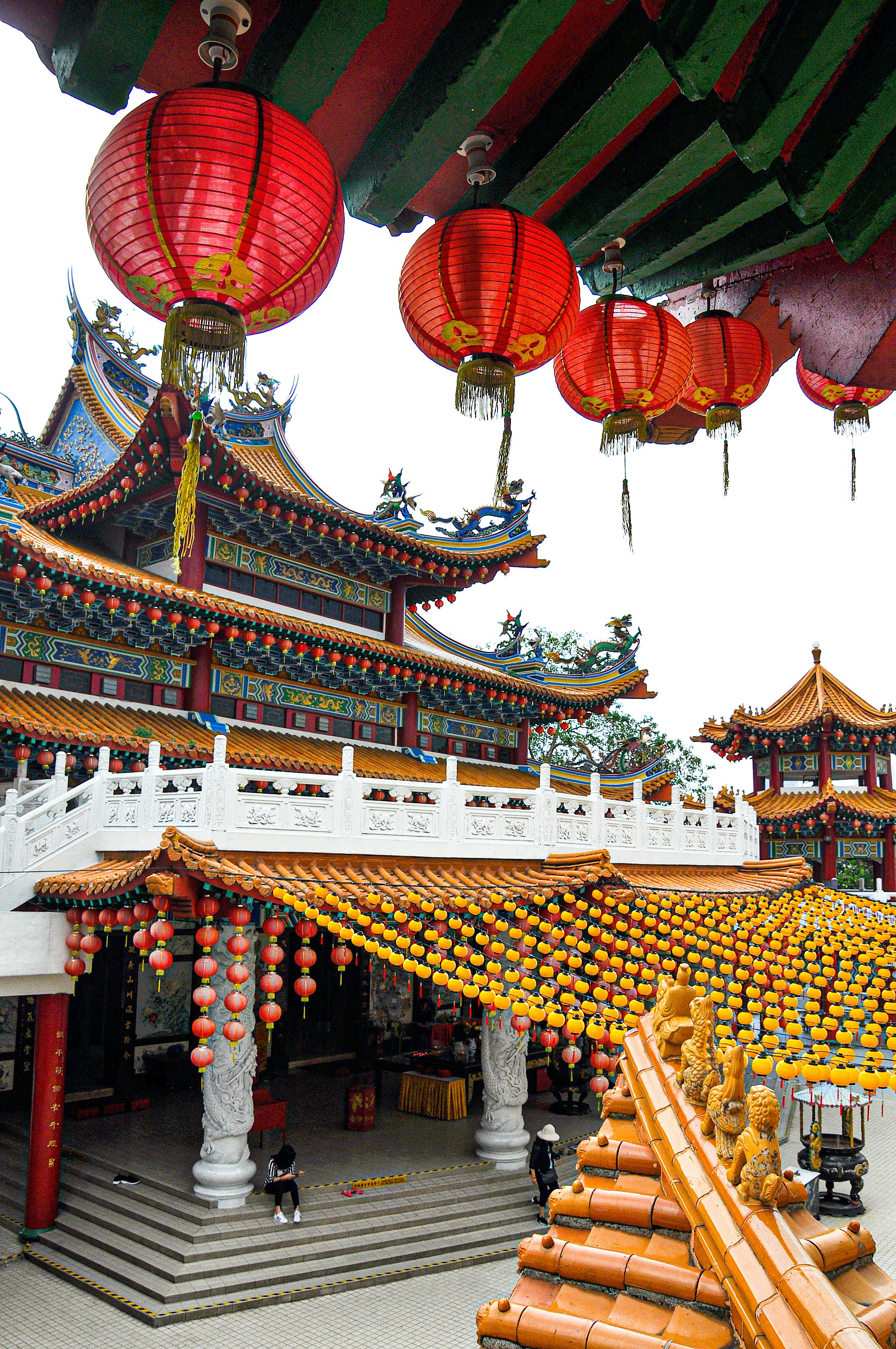 Vibrant red lanterns hang above intricate temple architecture adorned with colorful details and decorative elements. A bustling atmosphere reflects cultural heritage.