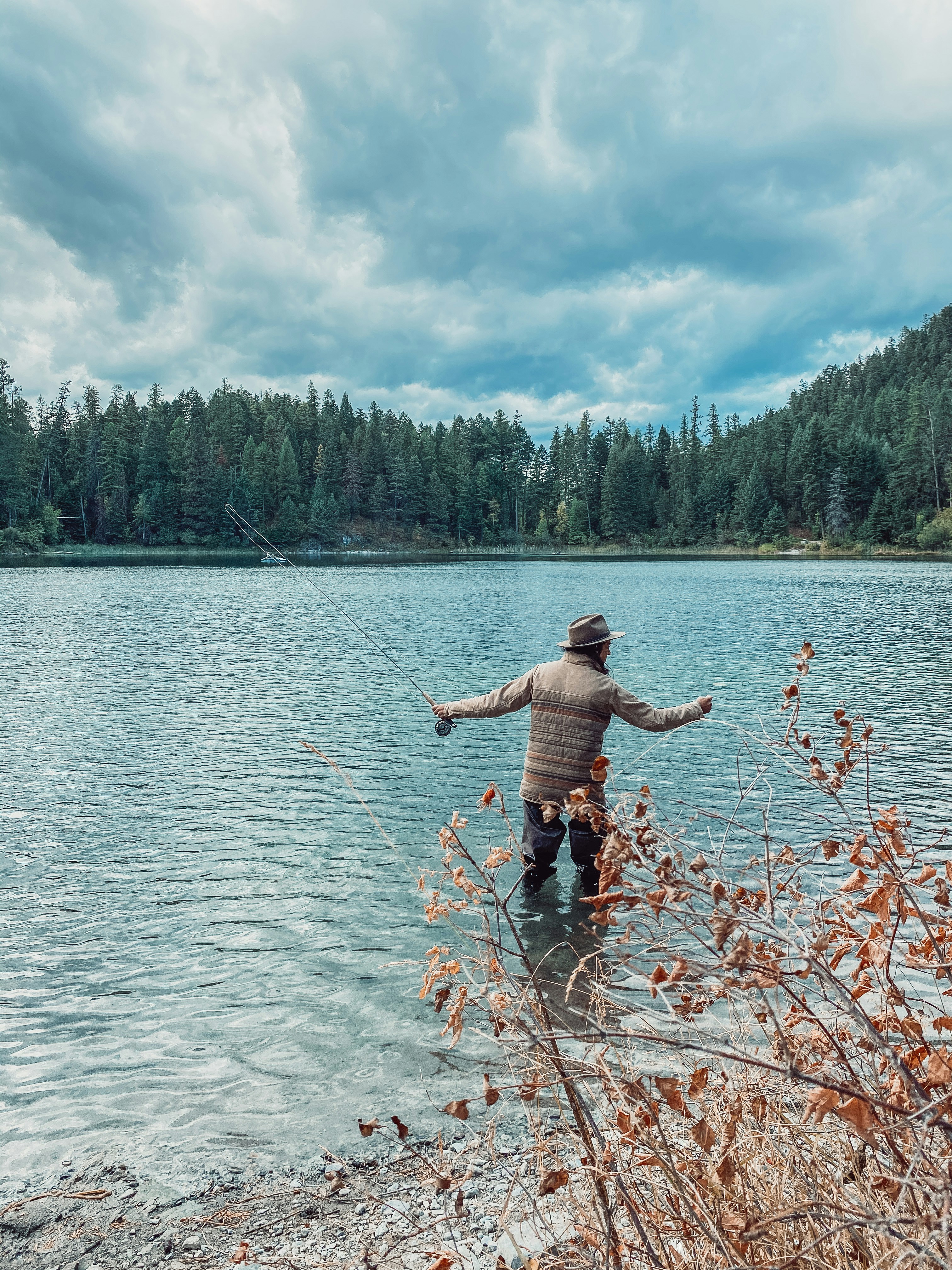A lone fisherman stands in a serene lake, casting his line amidst a backdrop of lush trees and a cloudy sky.