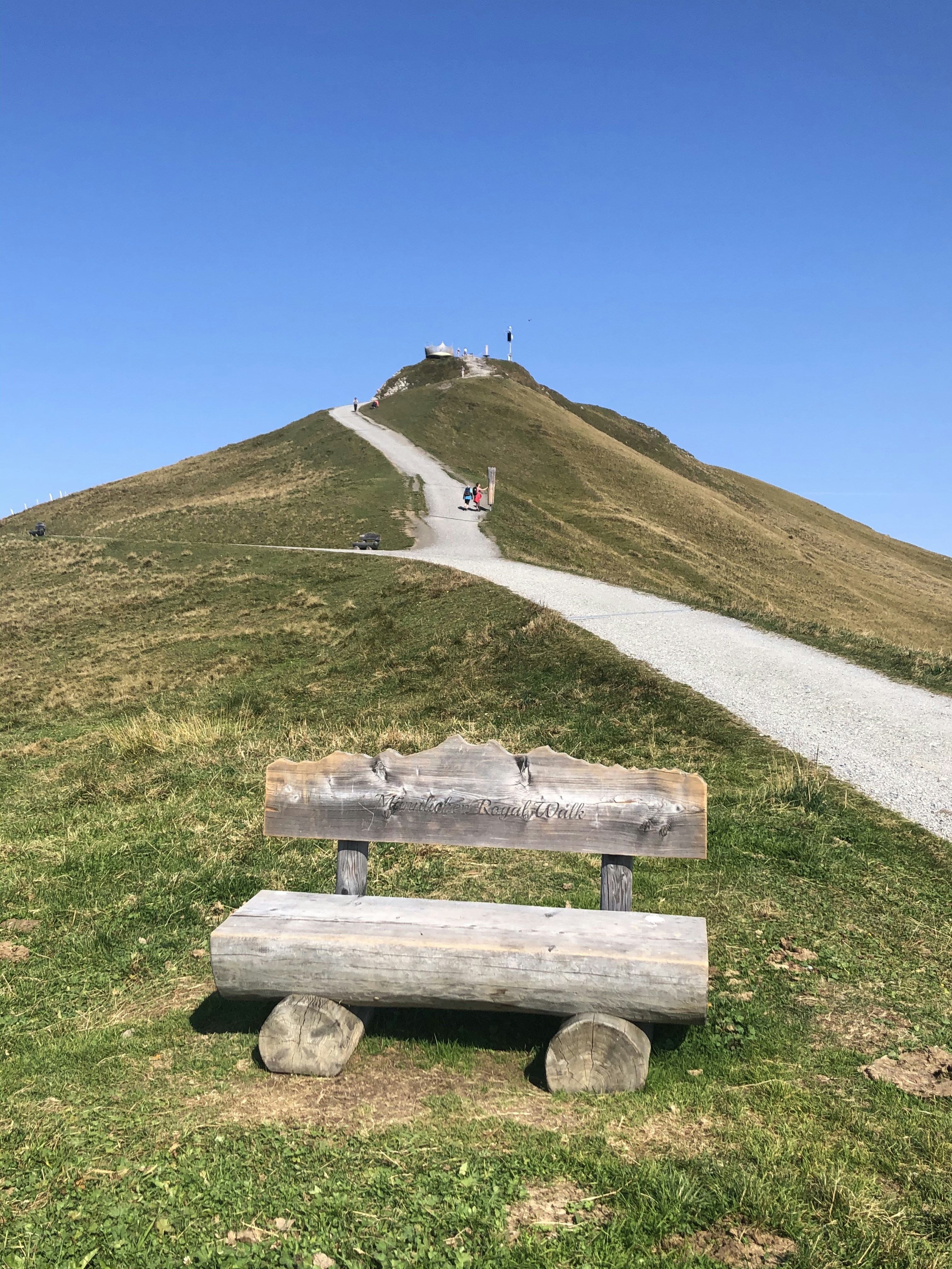 Wooden bench in the foreground with a winding path leading to a hilltop structure under a clear blue sky.