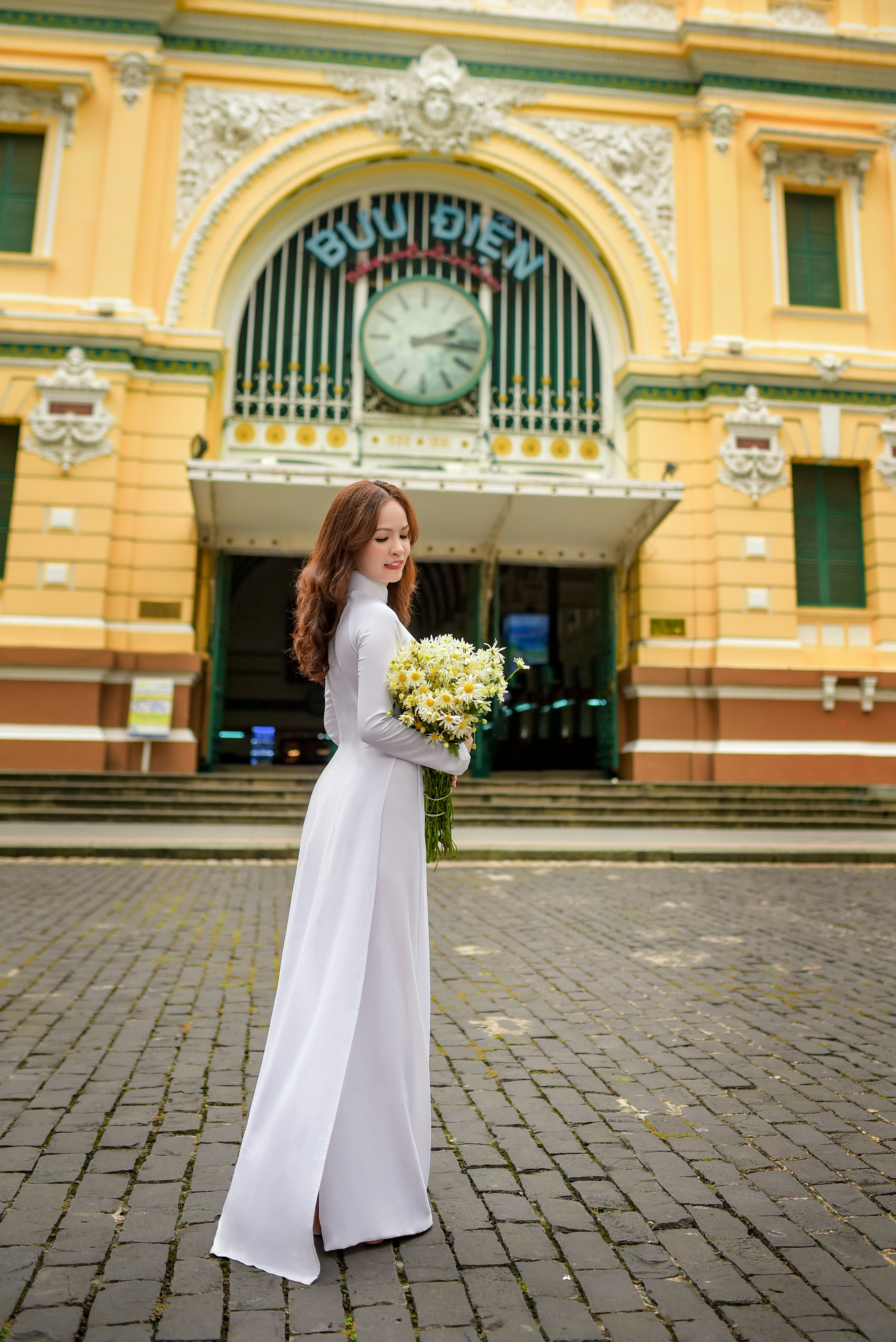 A woman in a flowing white dress stands gracefully with a bouquet of flowers in front of a historic building, showcasing architectural beauty and cultural heritage.