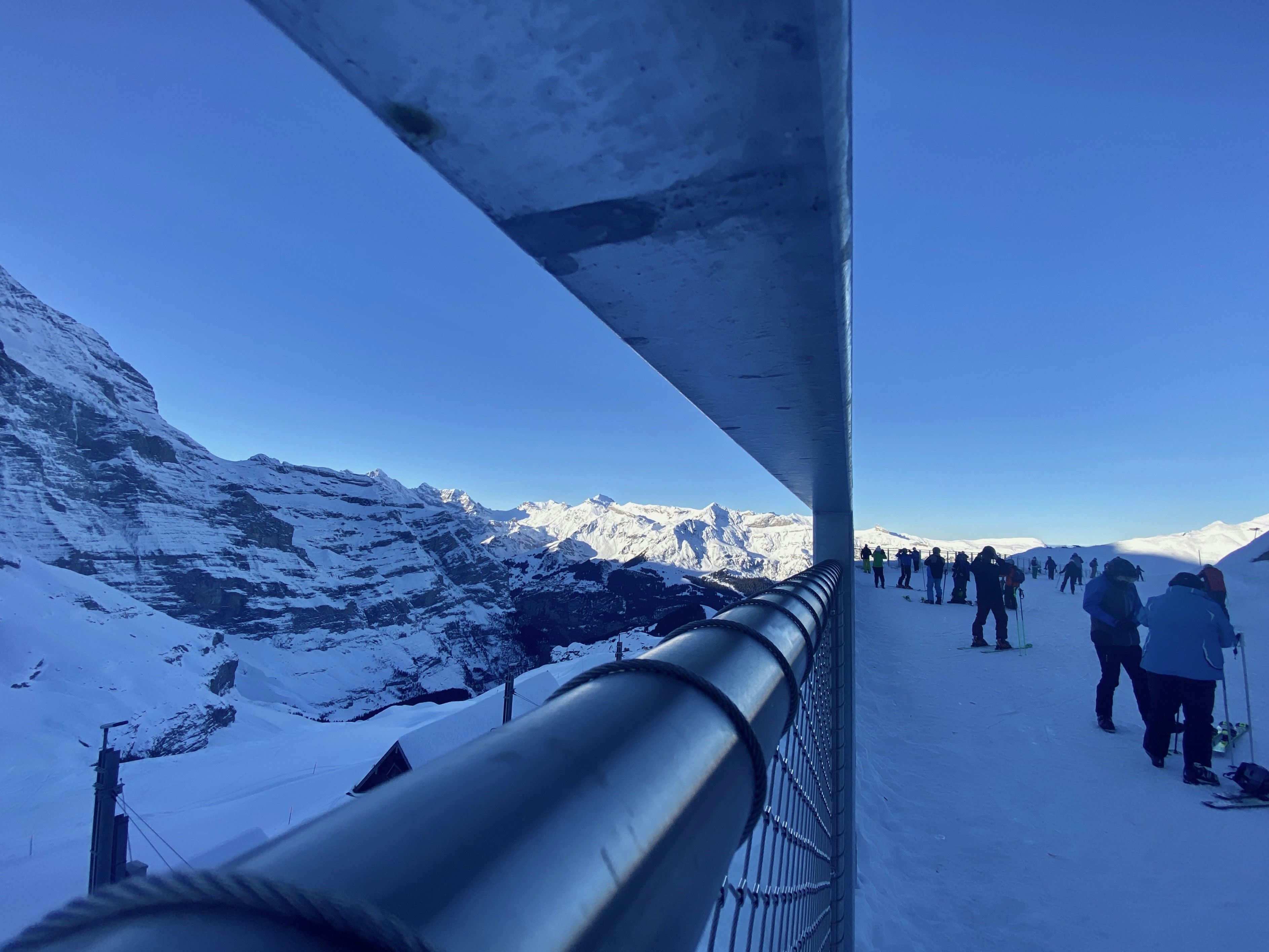 Skiers gather at a snowy overlook, framed by a metal railing, with majestic mountains in the background under a clear blue sky.